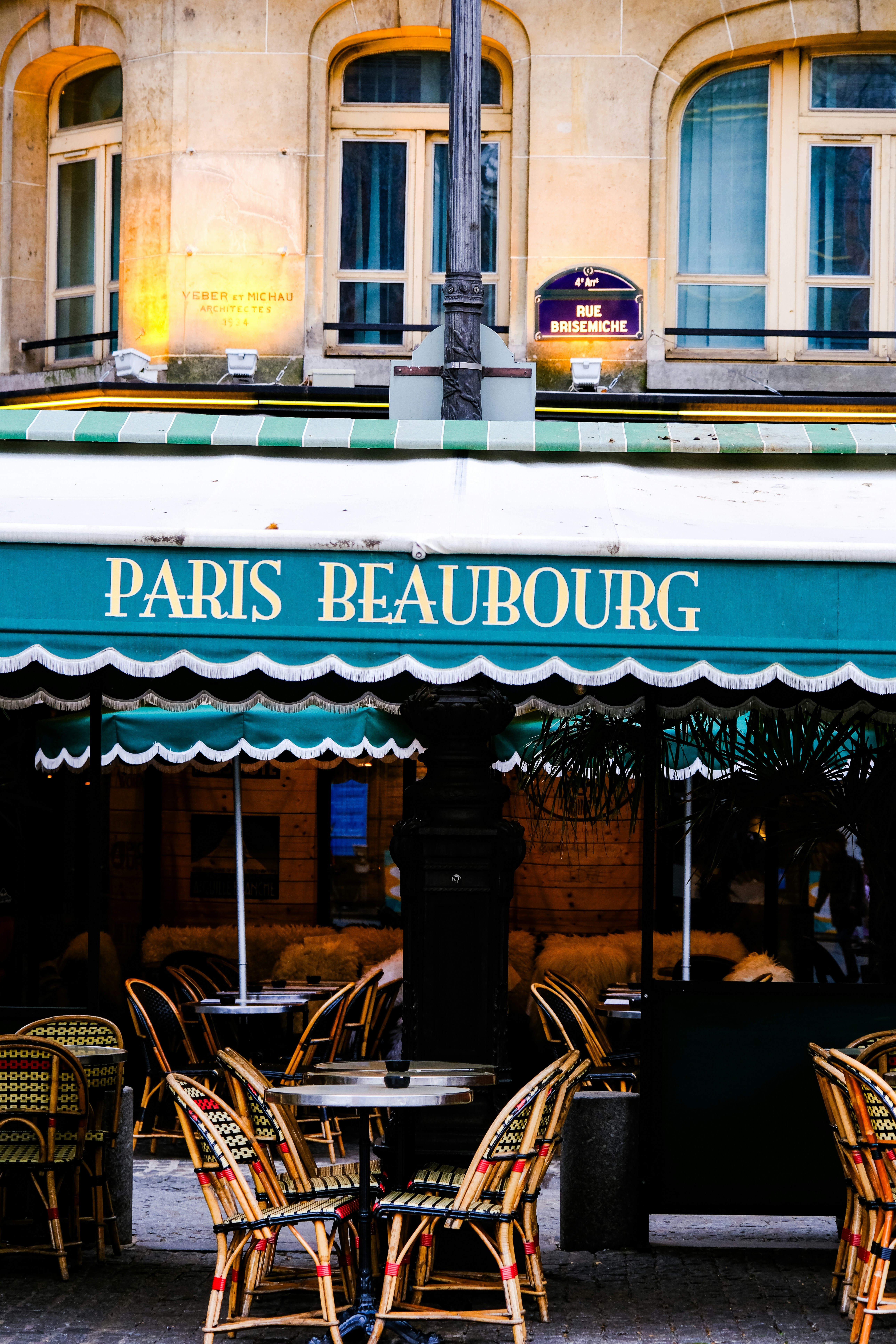 Outdoor café scene under a teal awning reading PARIS BEAUBOURG. Wicker chairs and round tables line the storefront with warm façades in the background.