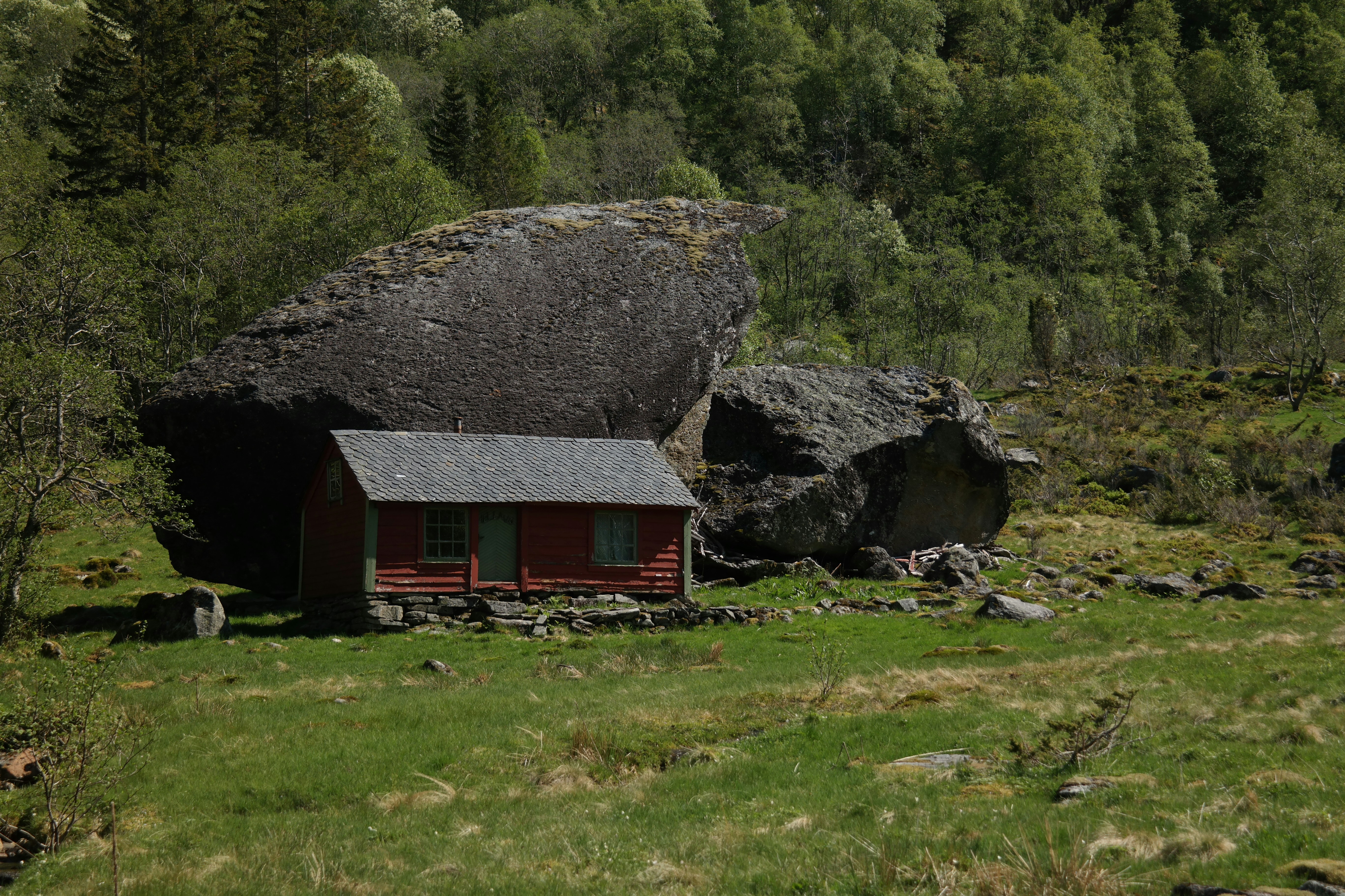 Red wooden cabin sits in a sunlit meadow beneath a massive dark boulder, with scattered rocks nearby. Tall trees form a dense green backdrop.