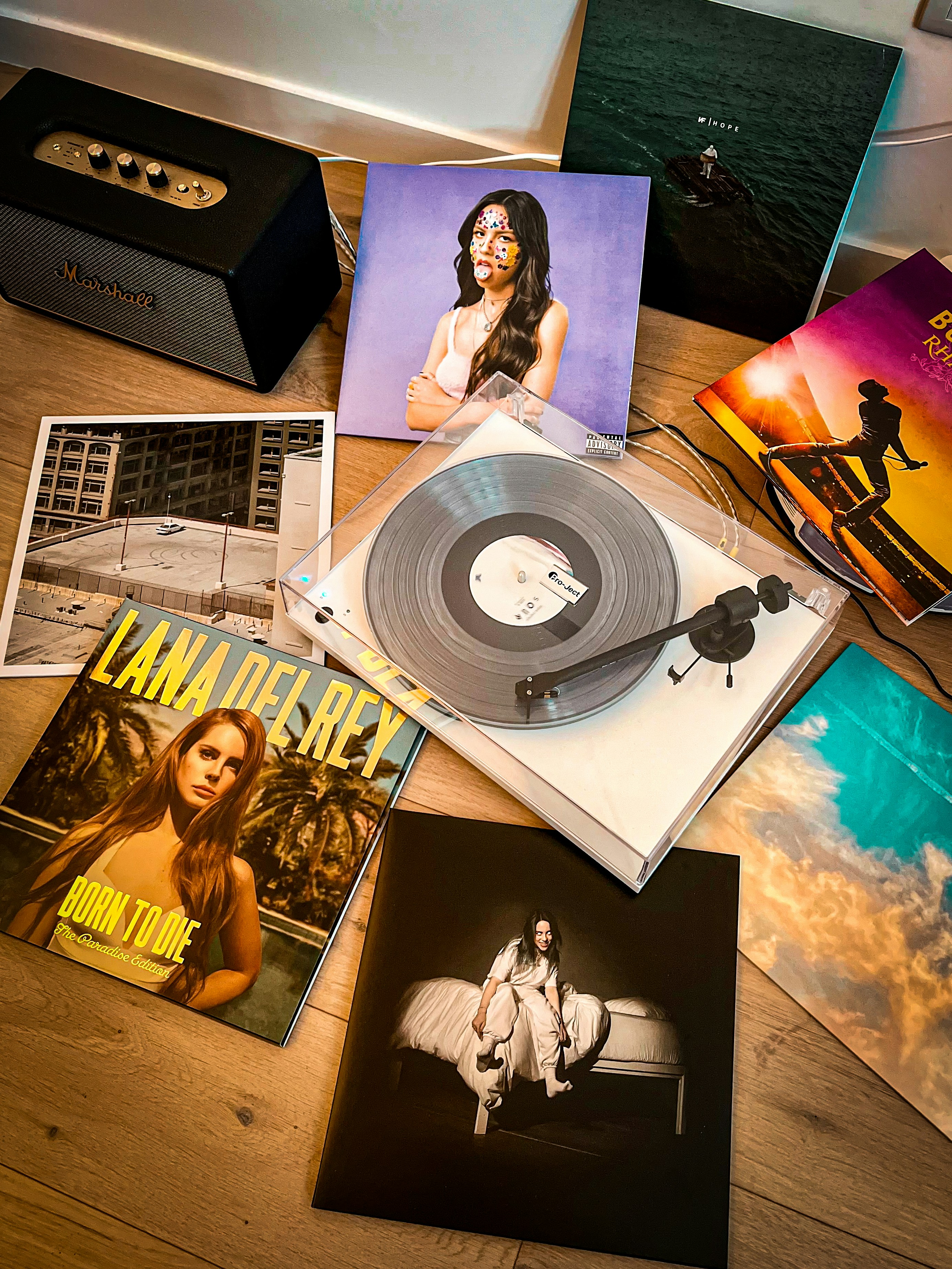 Turntable on a wooden floor surrounded by vinyl LPs and album covers, with a Marshall speaker visible in the background.