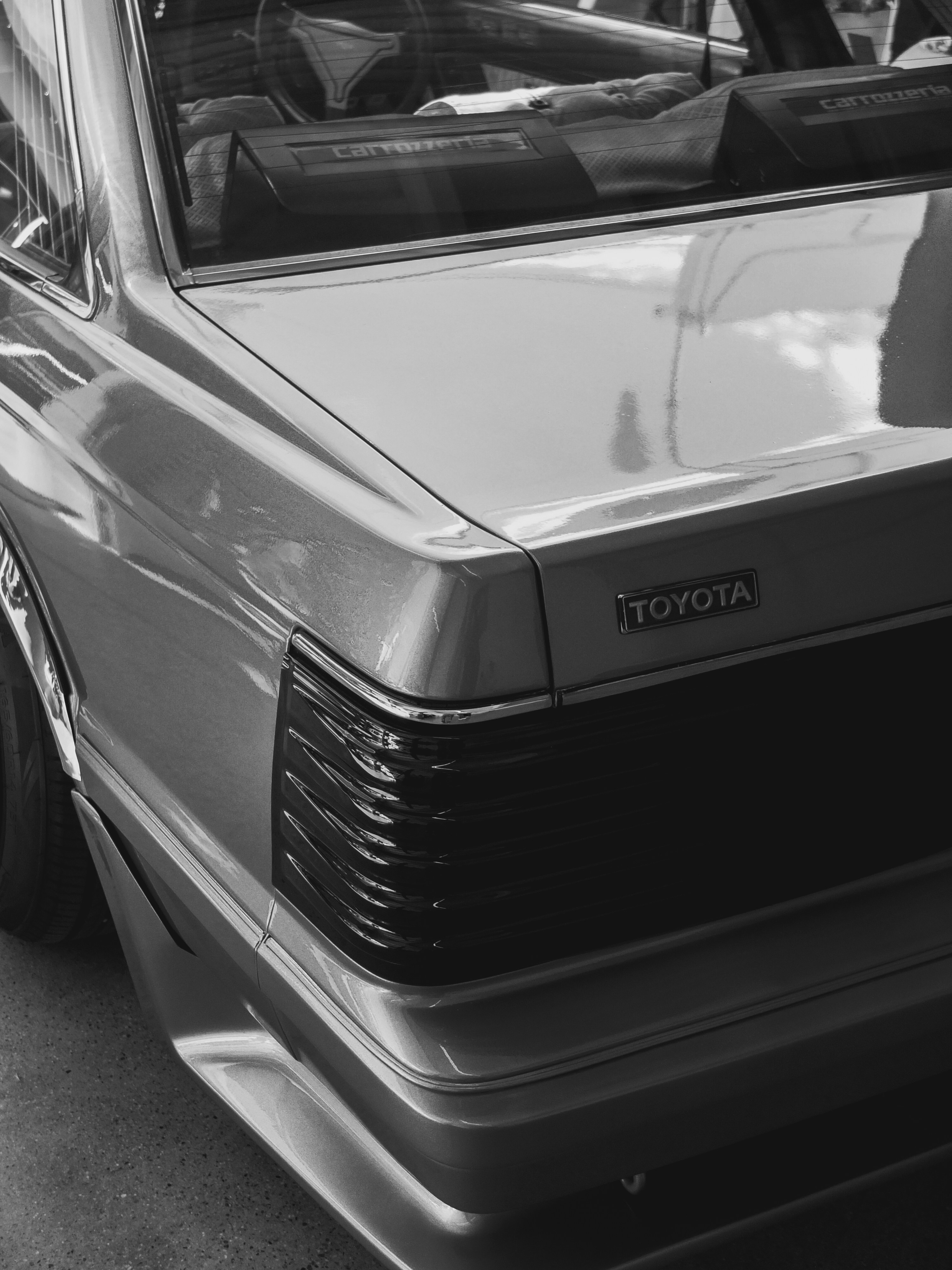 Black-and-white photograph of the rear end of a Toyota sedan, featuring the Toyota logo badge on the trunk. Glossy reflections emphasize the car's sculpted lines and taillight details.