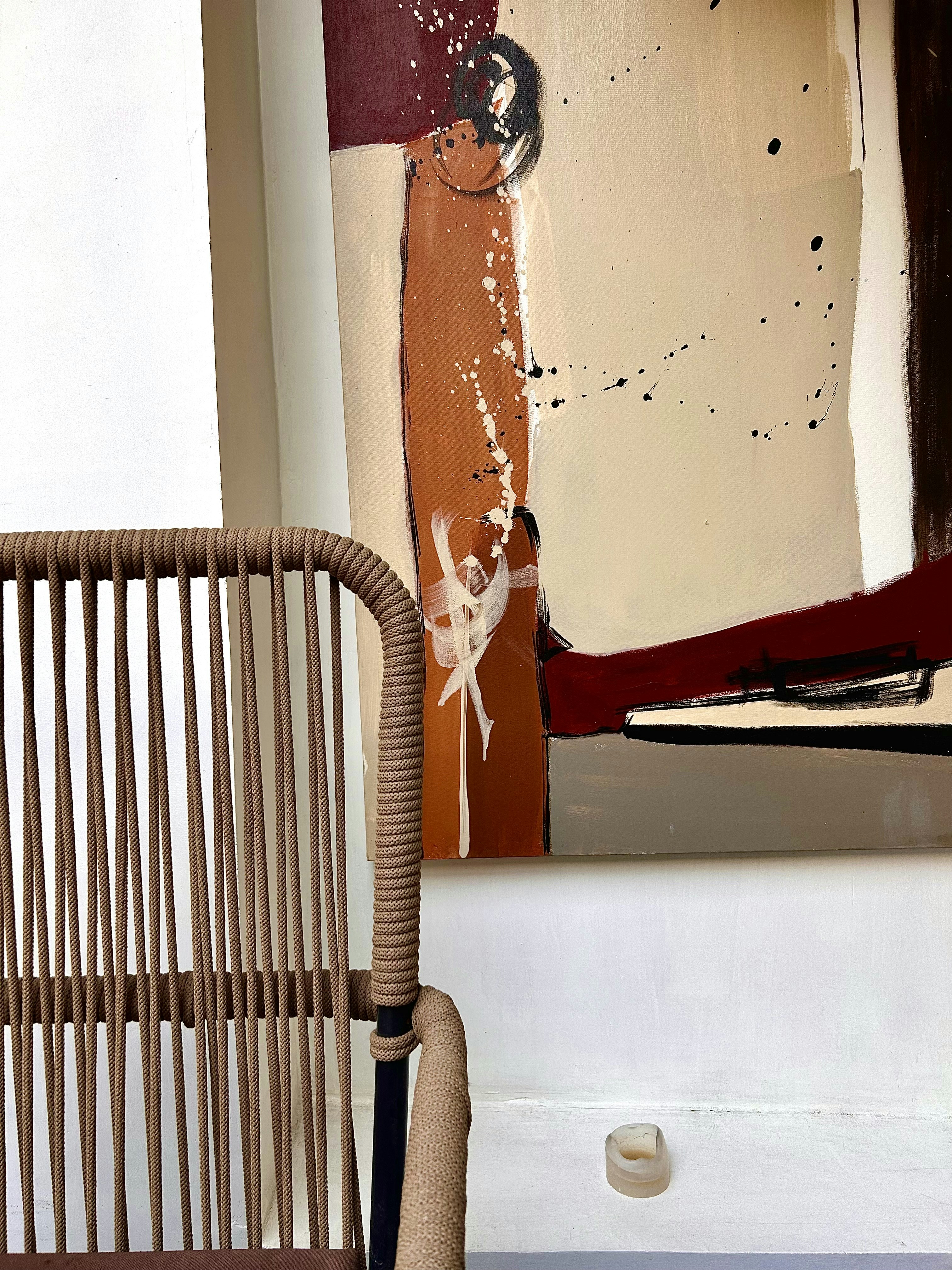 Abstract painting on a white wall with earthy blocks and a circular motif near the top. A woven chair foreground adds texture and depth, turning the scene into a quiet still-life study.