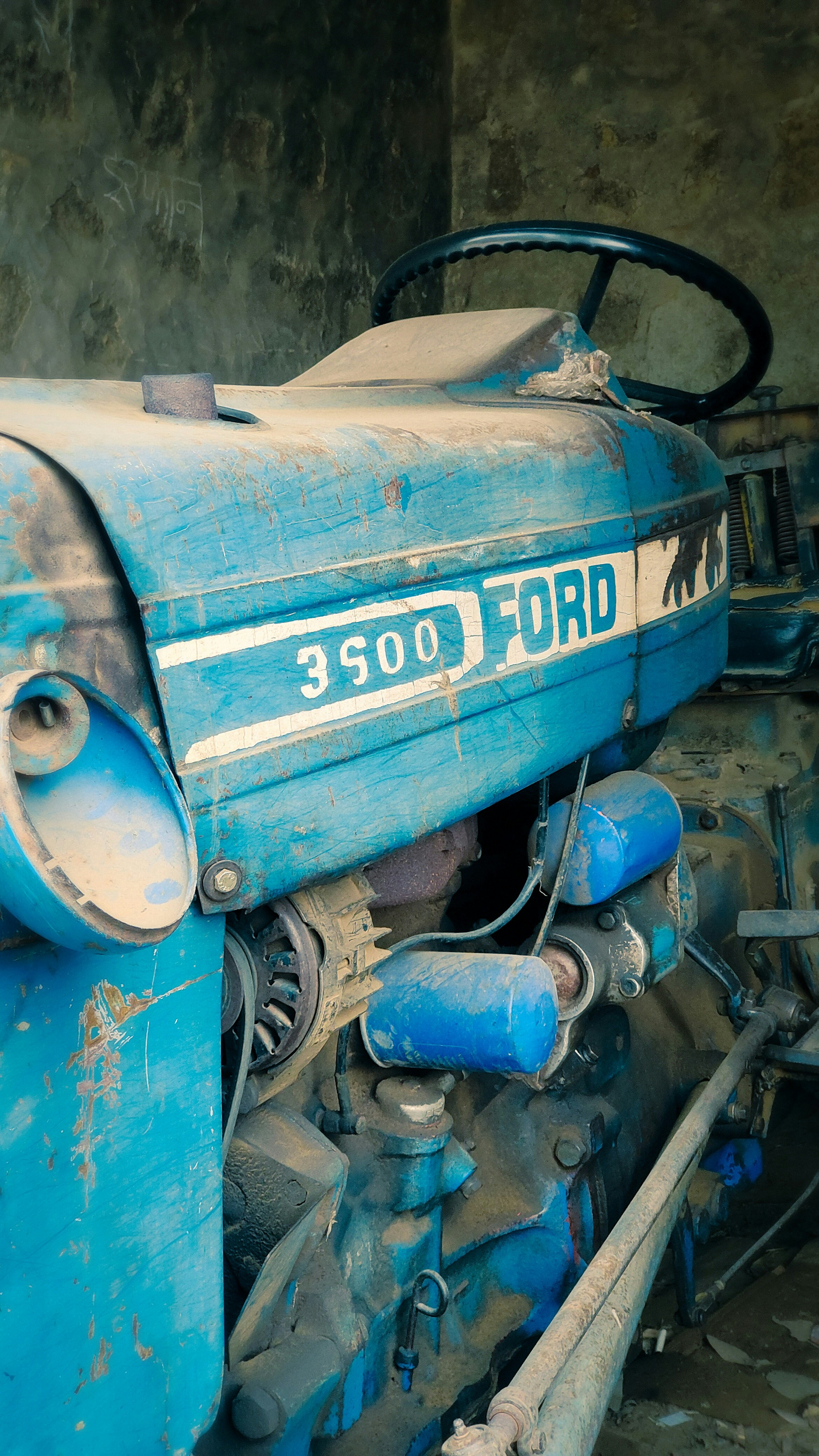 Weathered blue Ford 3500 tractor sits in a dusty, dim shed. The worn metal and exposed engine details emphasize its age.