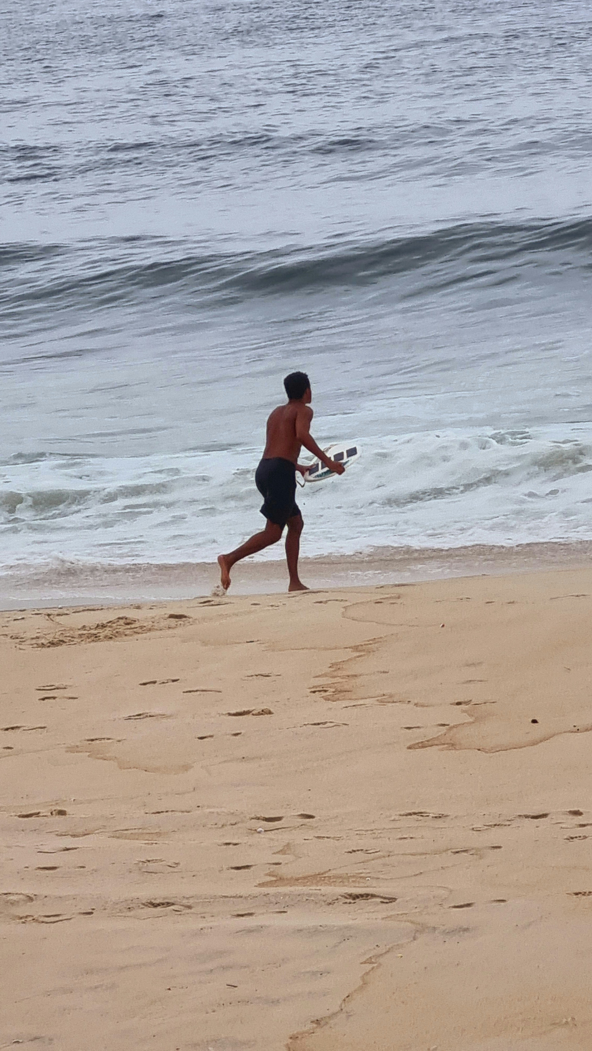 A man runs along the sandy beach toward the surf with a small kiteboard in hand, waves rolling in the background. The scene captures motion and coastal activity.