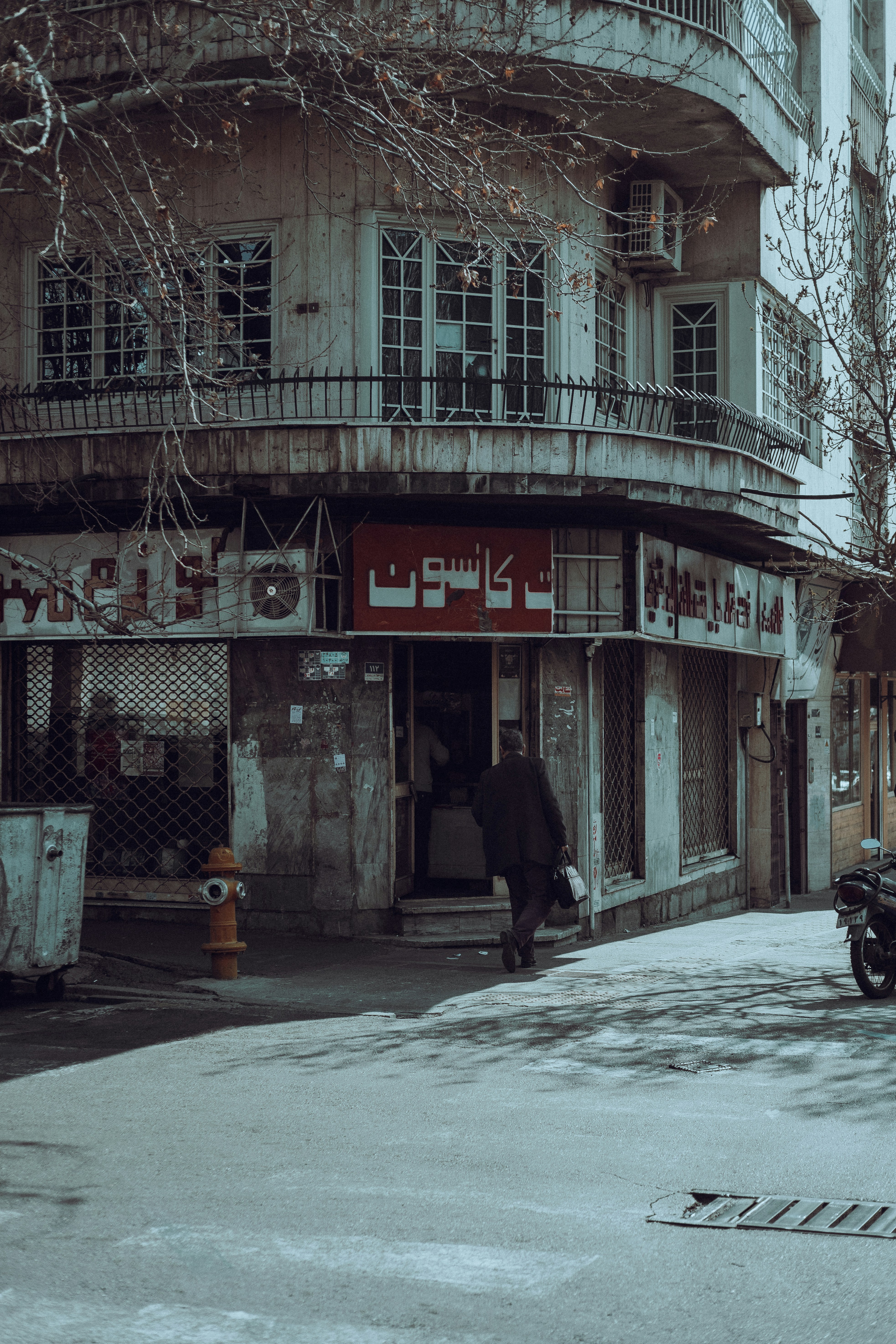 Snow at the Corner Shop: A man steps into a dim storefront beneath a weathered facade.