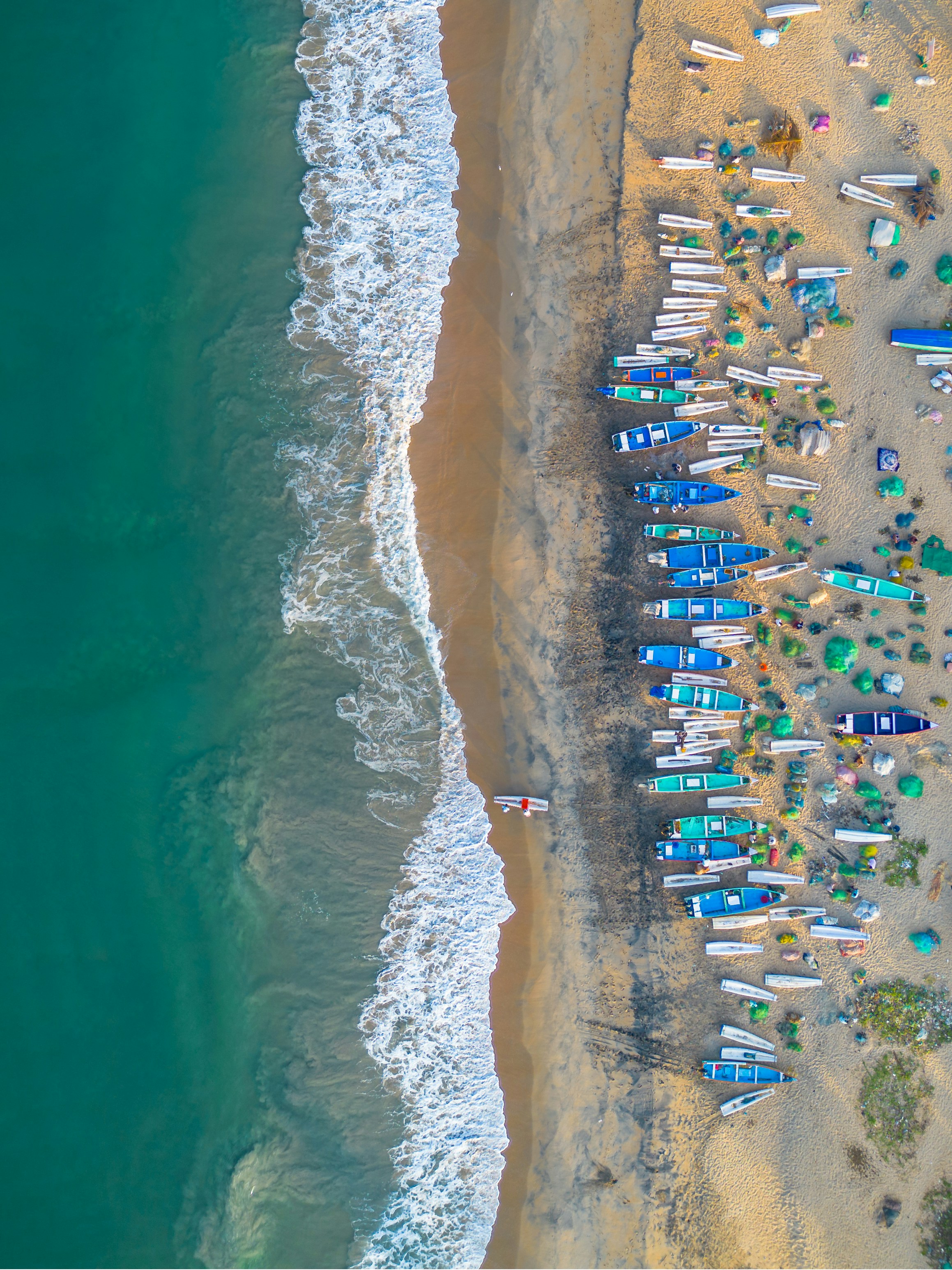 Aerial photograph showing a dense cluster of colorful boats along a sandy beach, with turquoise water on the left and foamy waves meeting the shore.