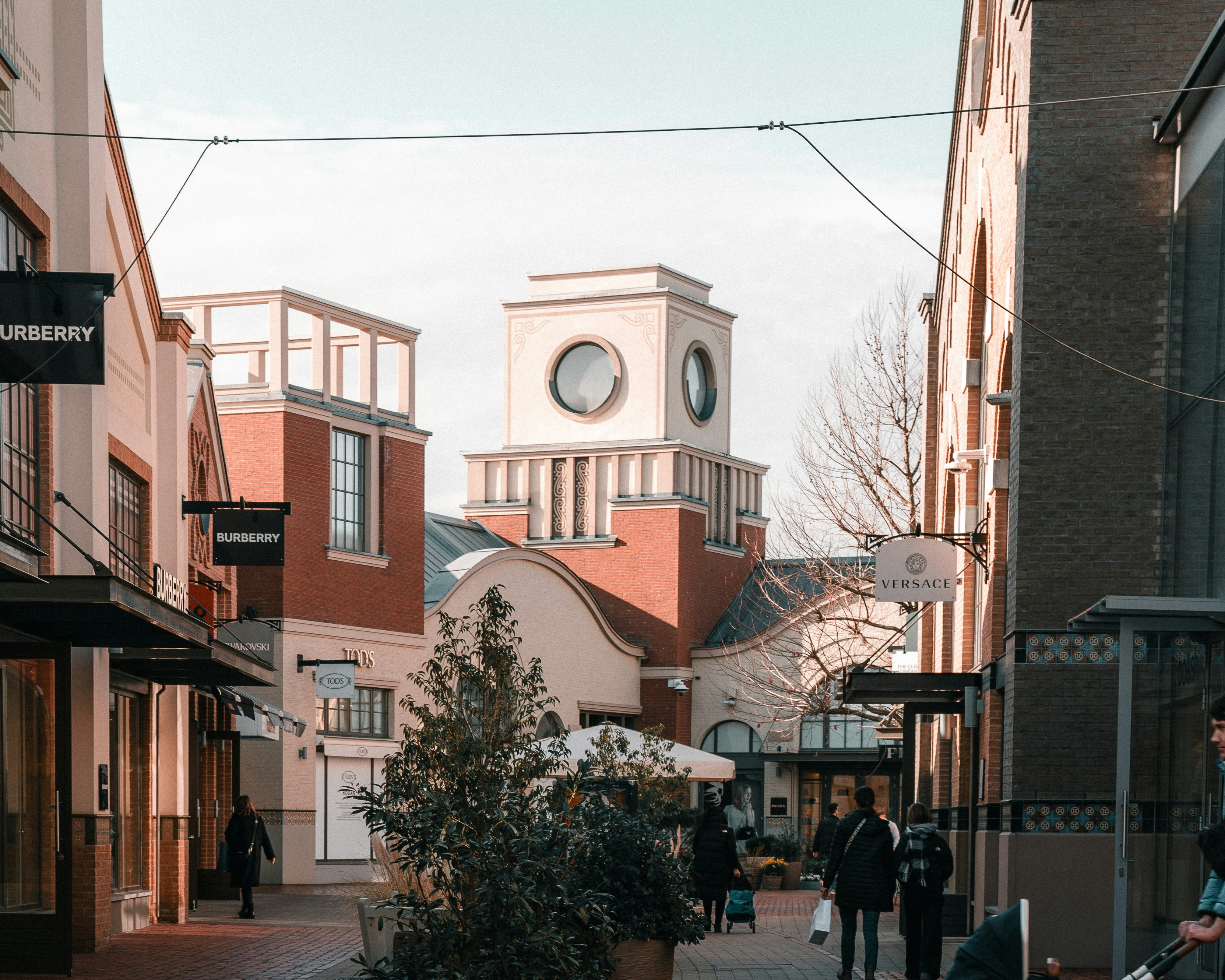 Street view of a quaint shopping area with a prominent clock tower and people strolling under a clear sky.