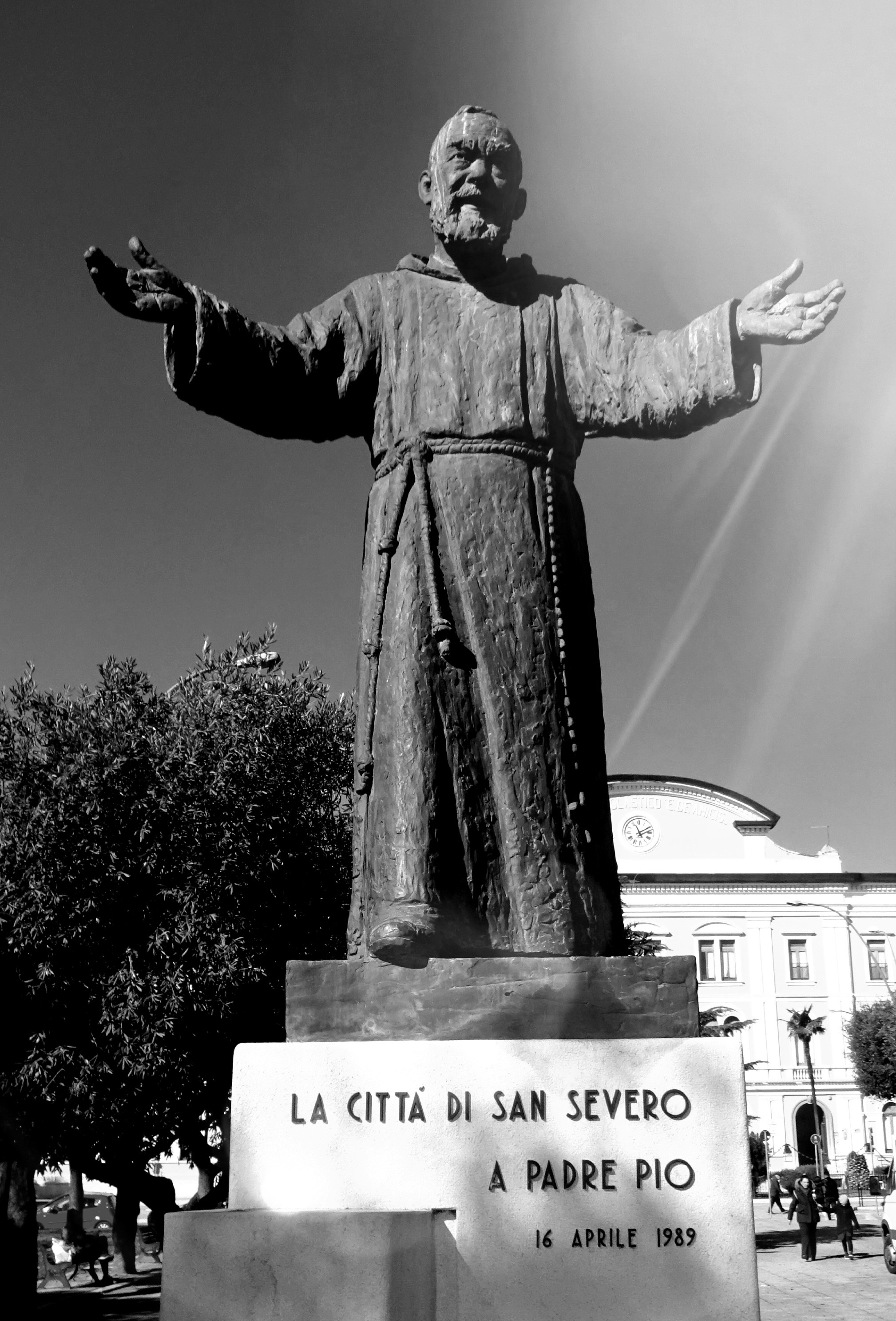 Black and white statue of a robed figure with outstretched arms in a public square under a bright sky.