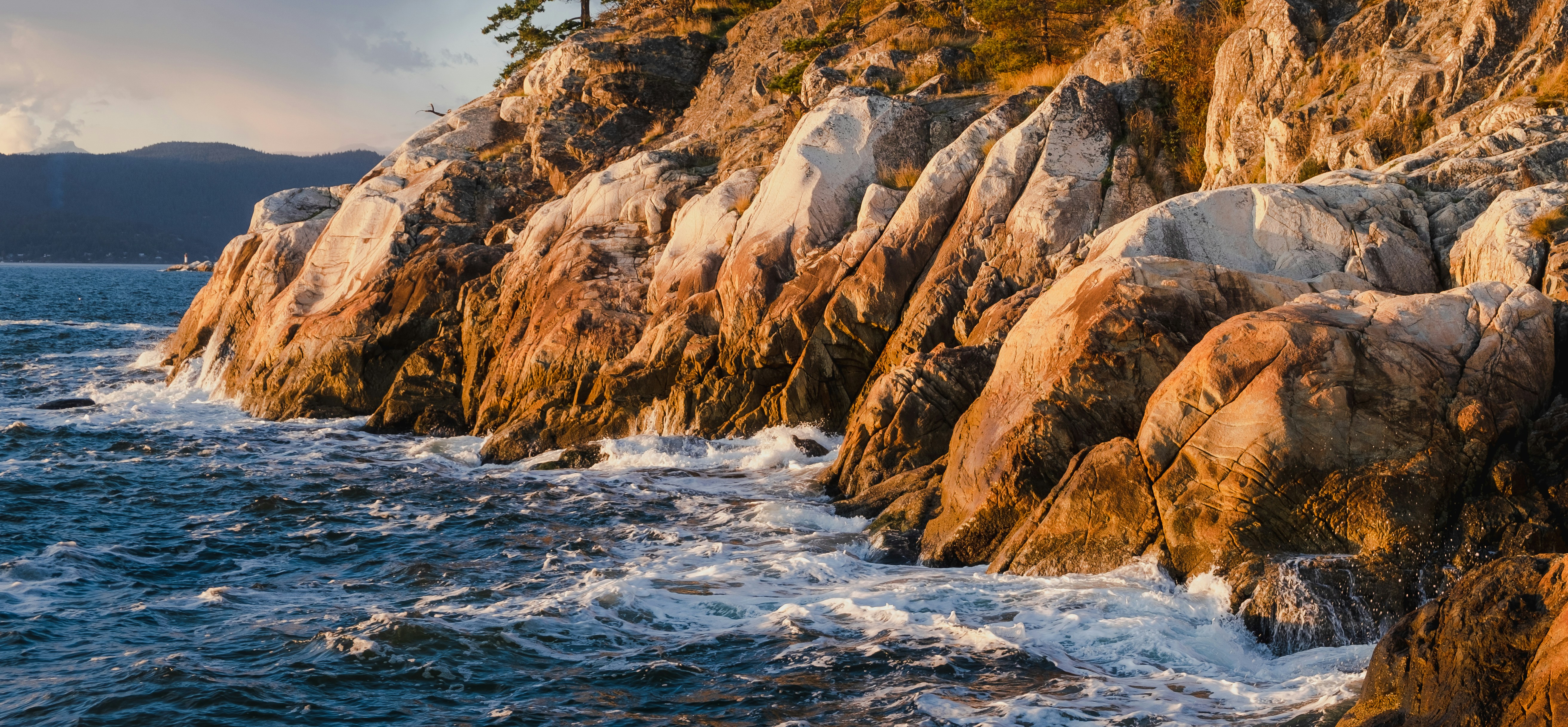 Waves crashing against rugged, sunlit cliffs on a coastal landscape.