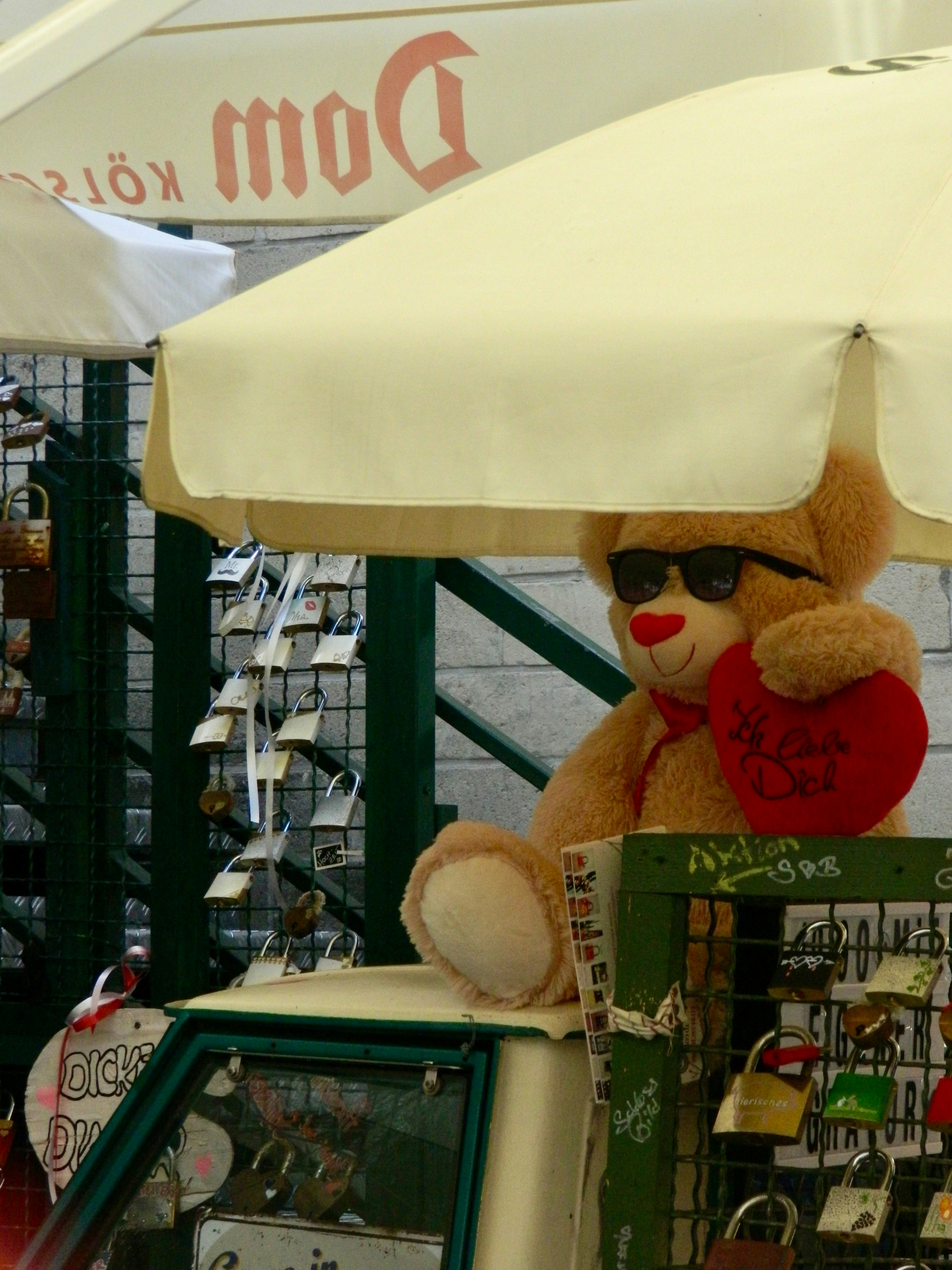 A plush teddy bear wearing sunglasses holds a heart-shaped sign, perched under a market umbrella amidst a backdrop of colorful locks and trinkets.