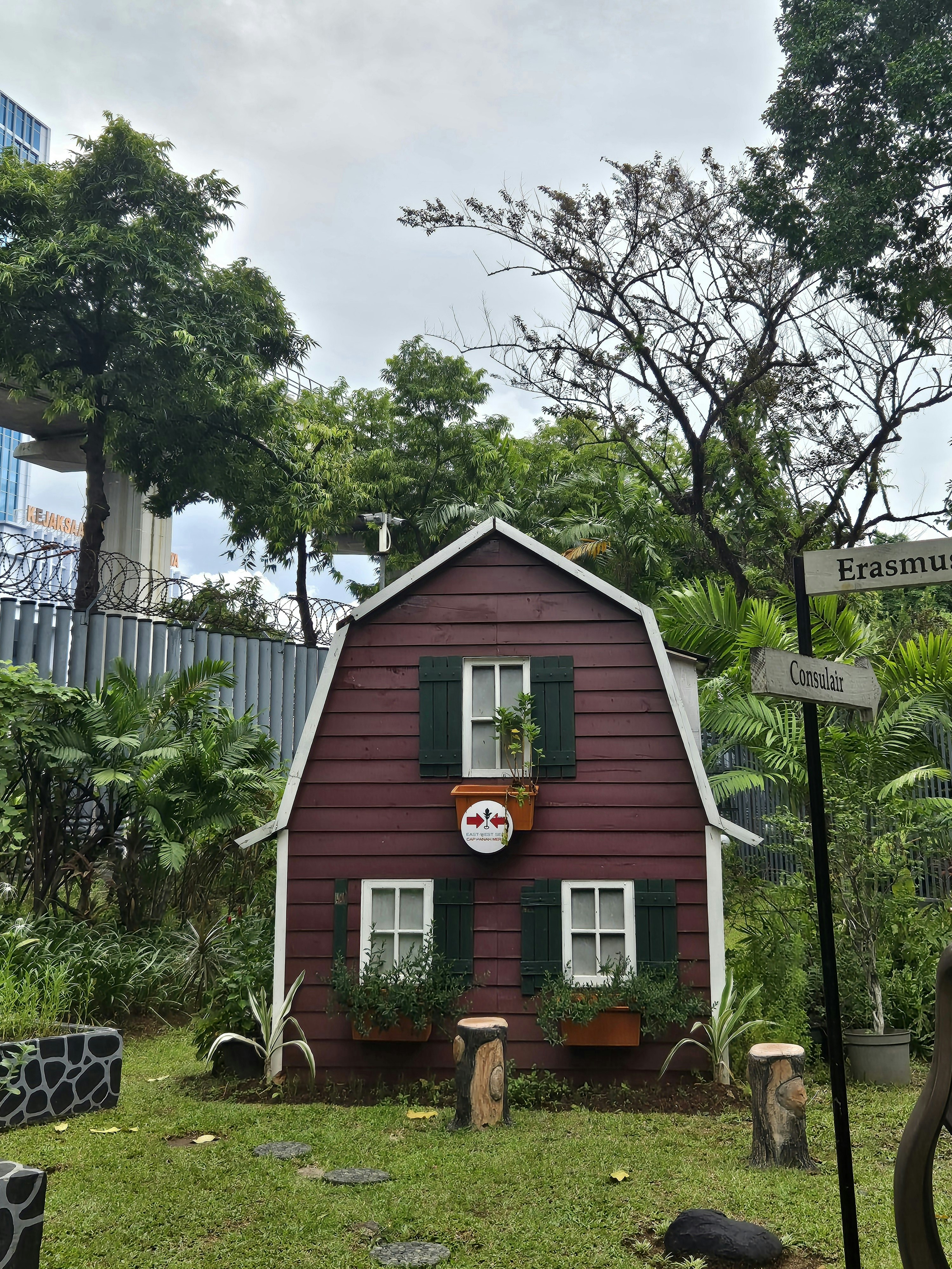 A photo of a miniature burgundy cottage with white trim and green shutters in a grassy yard, surrounded by plants and a signpost. The scene captures a quiet garden moment with natural daylight.