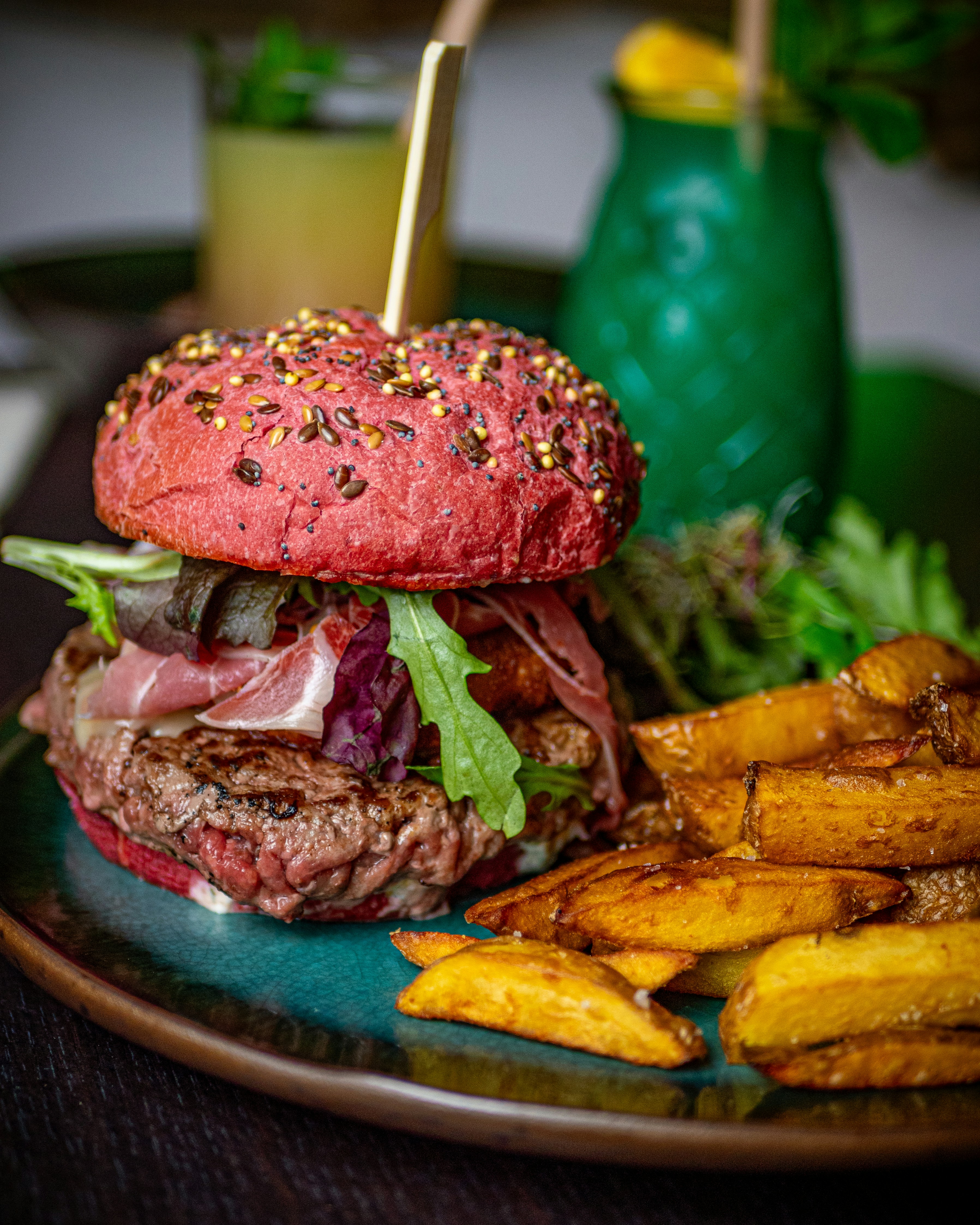Vibrant red burger topped with fresh greens and layers of meat, accompanied by golden potato wedges and a colorful drink in the background.