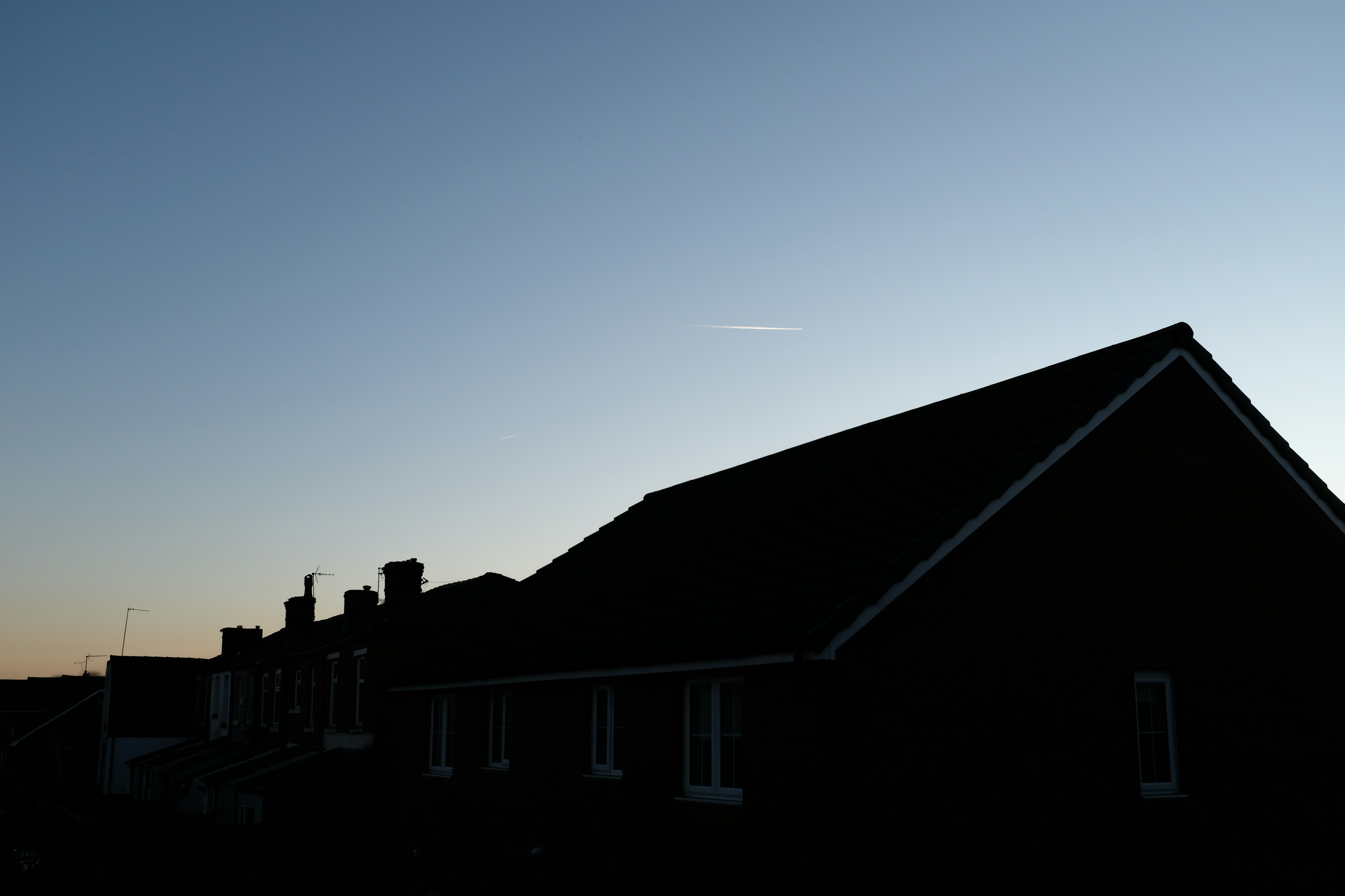 Contrail over a Quiet Dusk: Silhouetted Rooftops Against a Pale Sky