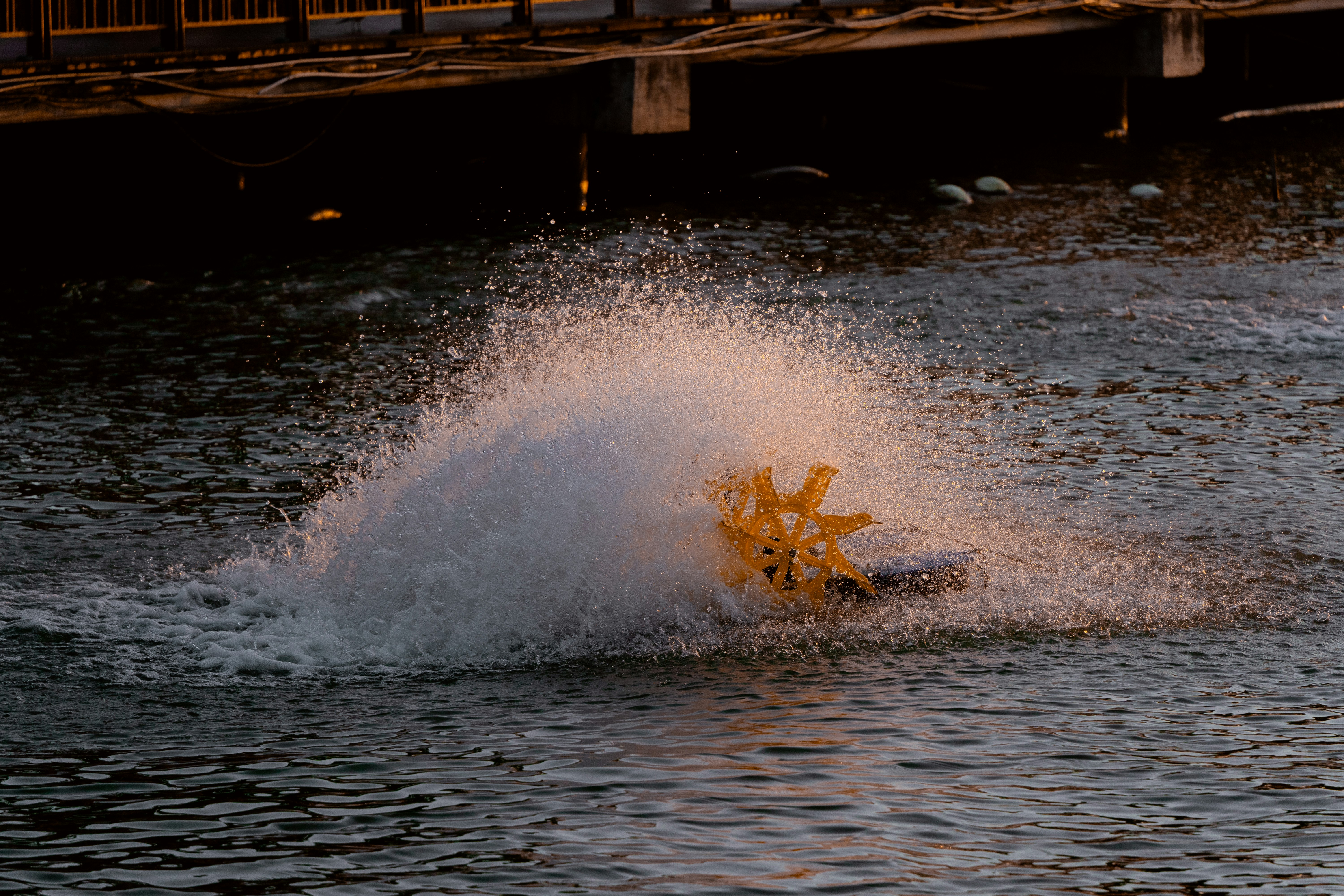 Orange paddle wheel creating a large splash in a calm river under a wooden bridge.