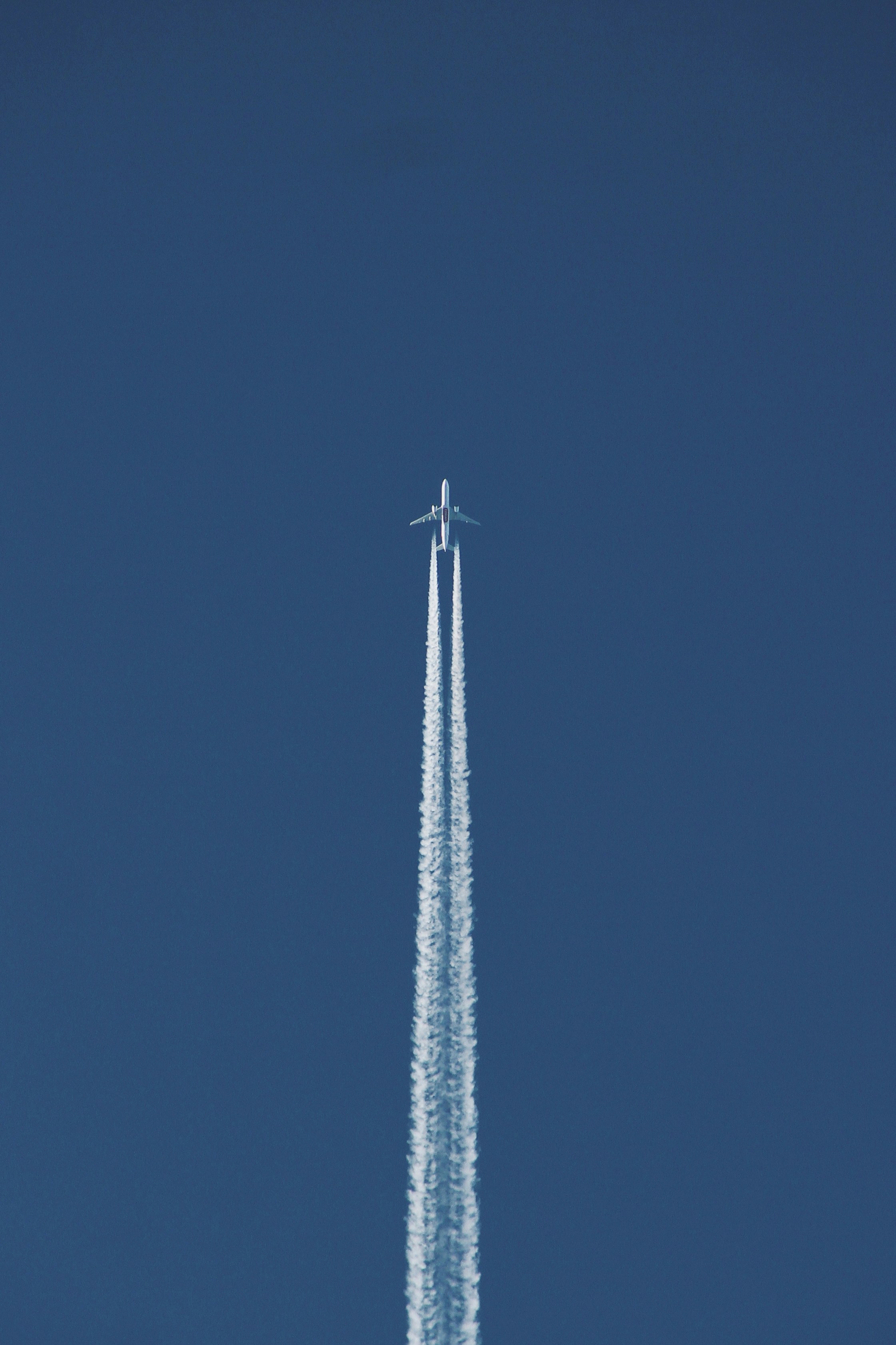 Airplane soaring high above, leaving behind two distinct contrails against a clear blue sky.