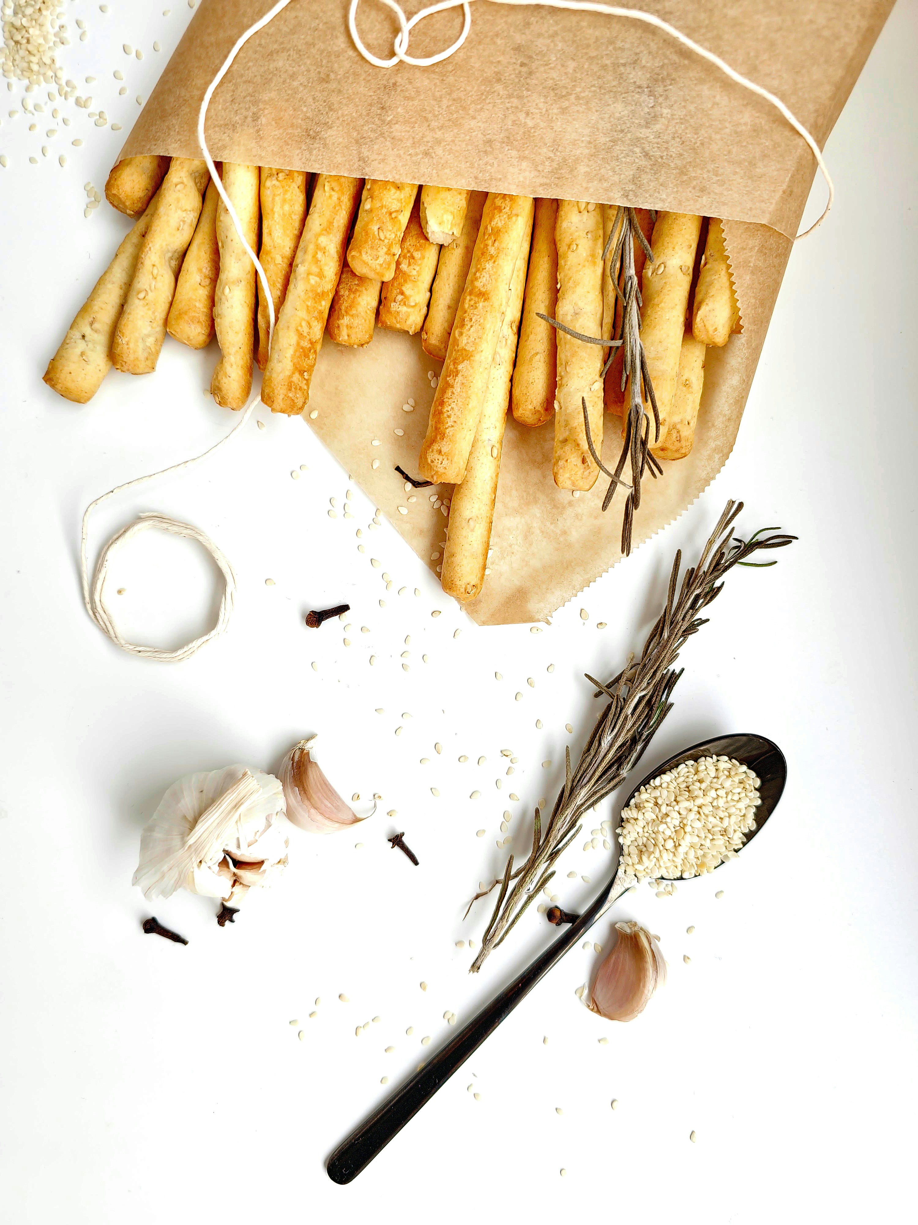 A rustic bundle of golden breadsticks nestled in parchment, accompanied by garlic cloves, rosemary sprigs, and sesame seeds on a white backdrop.