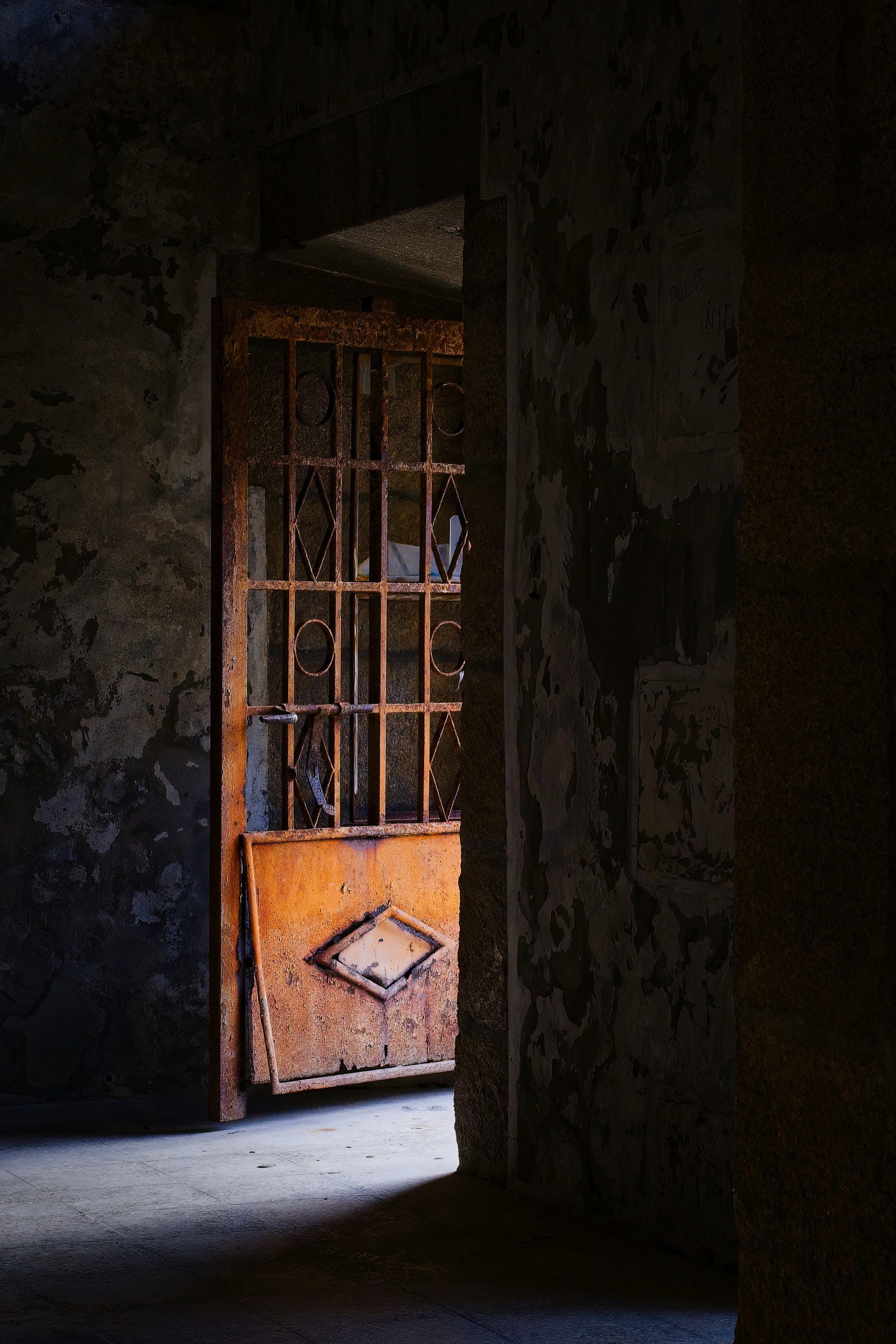 A Glimpse Through an Old, Rusty Doorway in a Dark, Abandoned Building - A narrow doorway reveals a glimpse of a room in a dark, abandoned building. The rusty, orange door is partially open, allowing a ray of light to illuminate the dusty, concrete floor. The walls are cracked and weathered, suggesting the building has stood empty for many years.