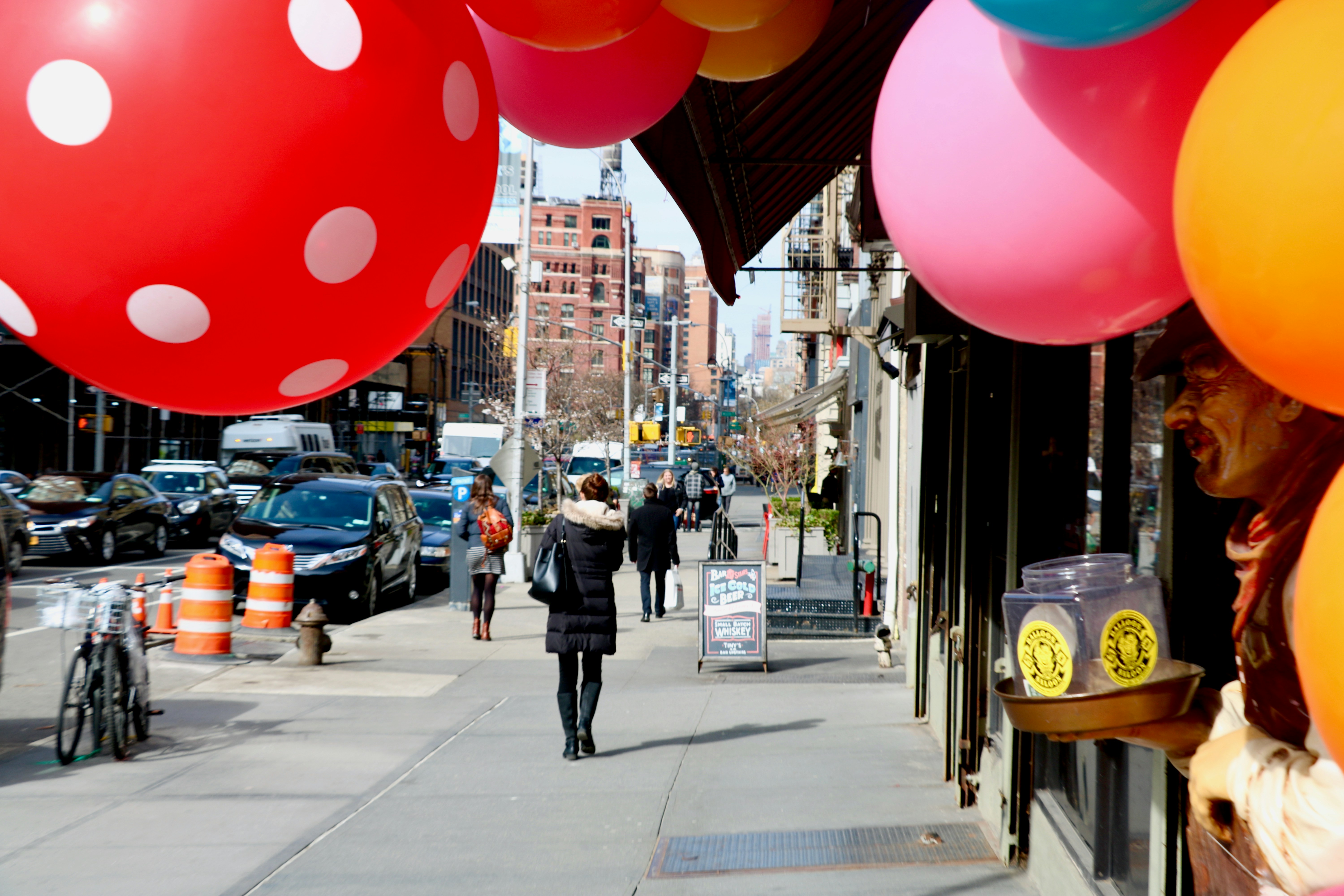 A lady walks on the sidewalk on a street of Lower Manhattan past some colorful balloons