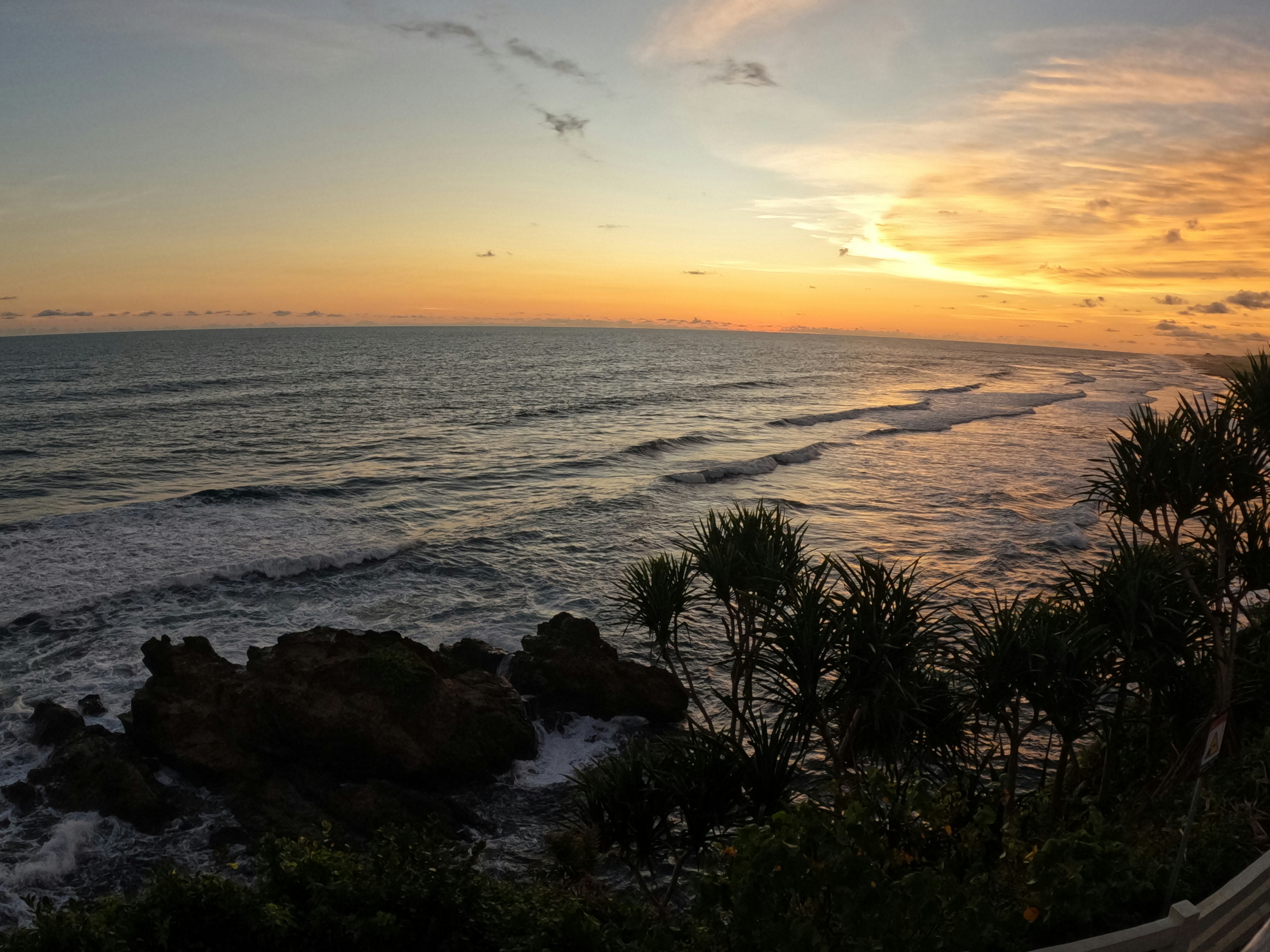 Sunset over the ocean with silhouetted coastal vegetation and distant clouds in Karangtawulan, Indonesia.