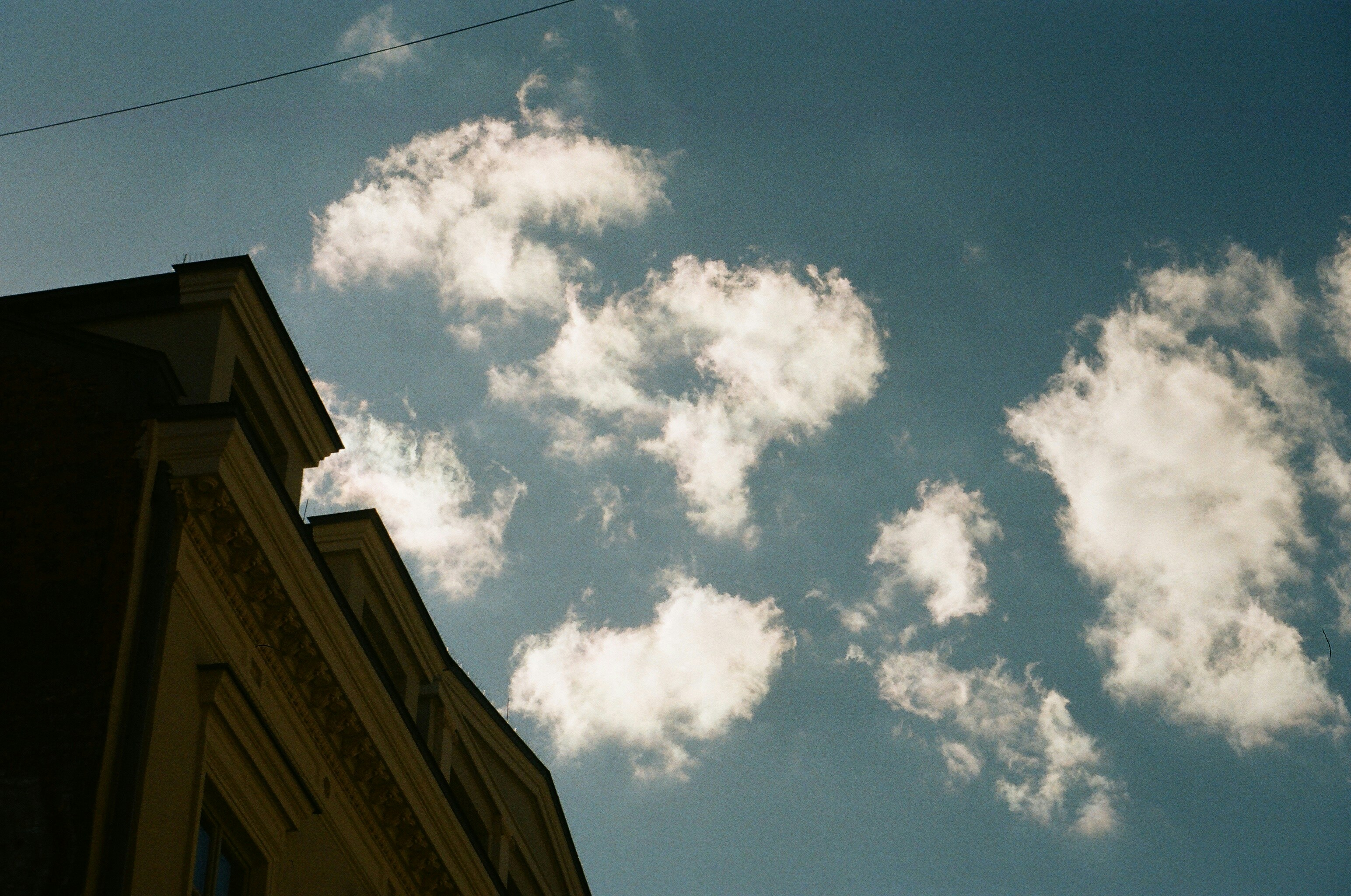 Low-angle shot of a sunlit building corner against a vast blue sky dotted with clouds. The image emphasizes architectural detail against an expansive atmospheric backdrop.