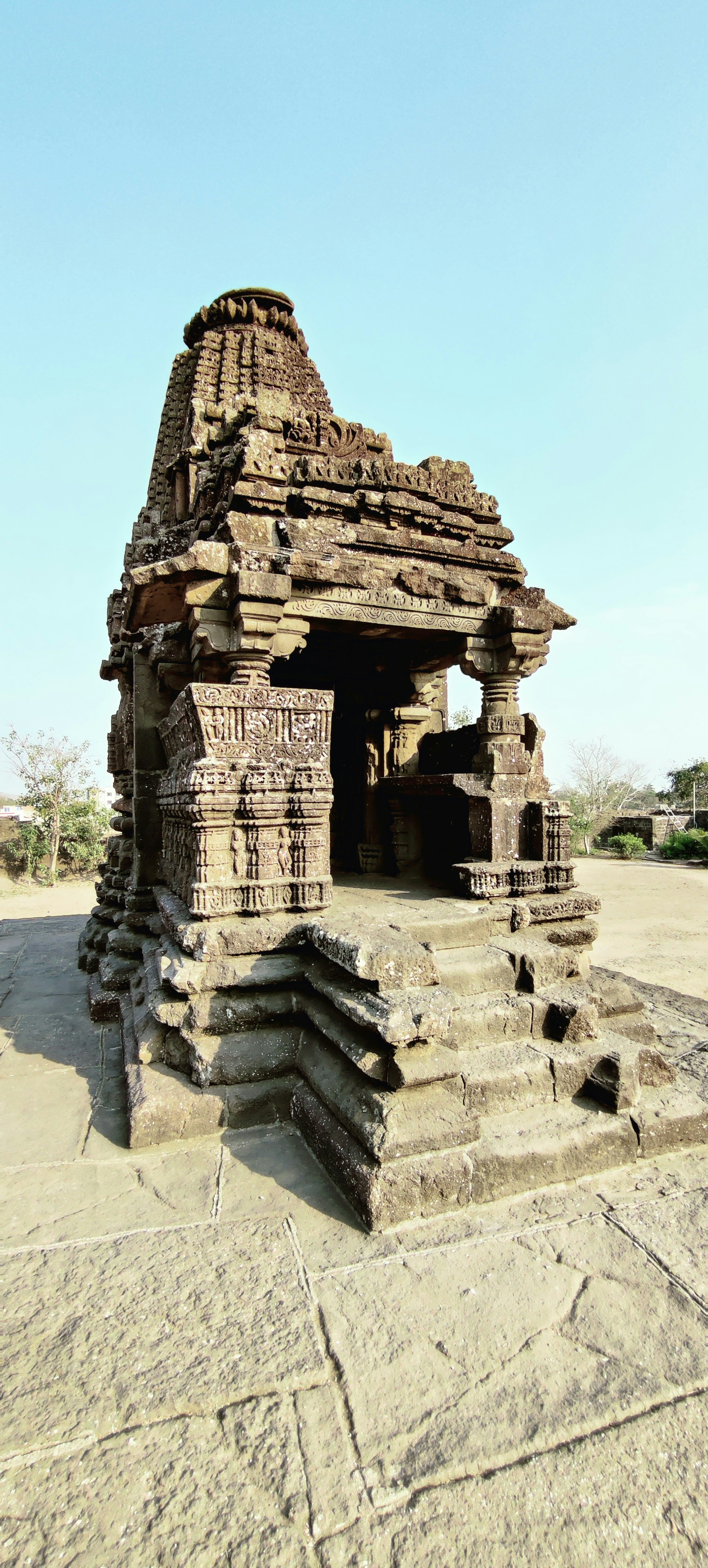 Weathered stone temple with ornate carvings sits on a stepped platform under a bright blue sky. The scene emphasizes ancient craftsmanship and the temple's solitary presence.