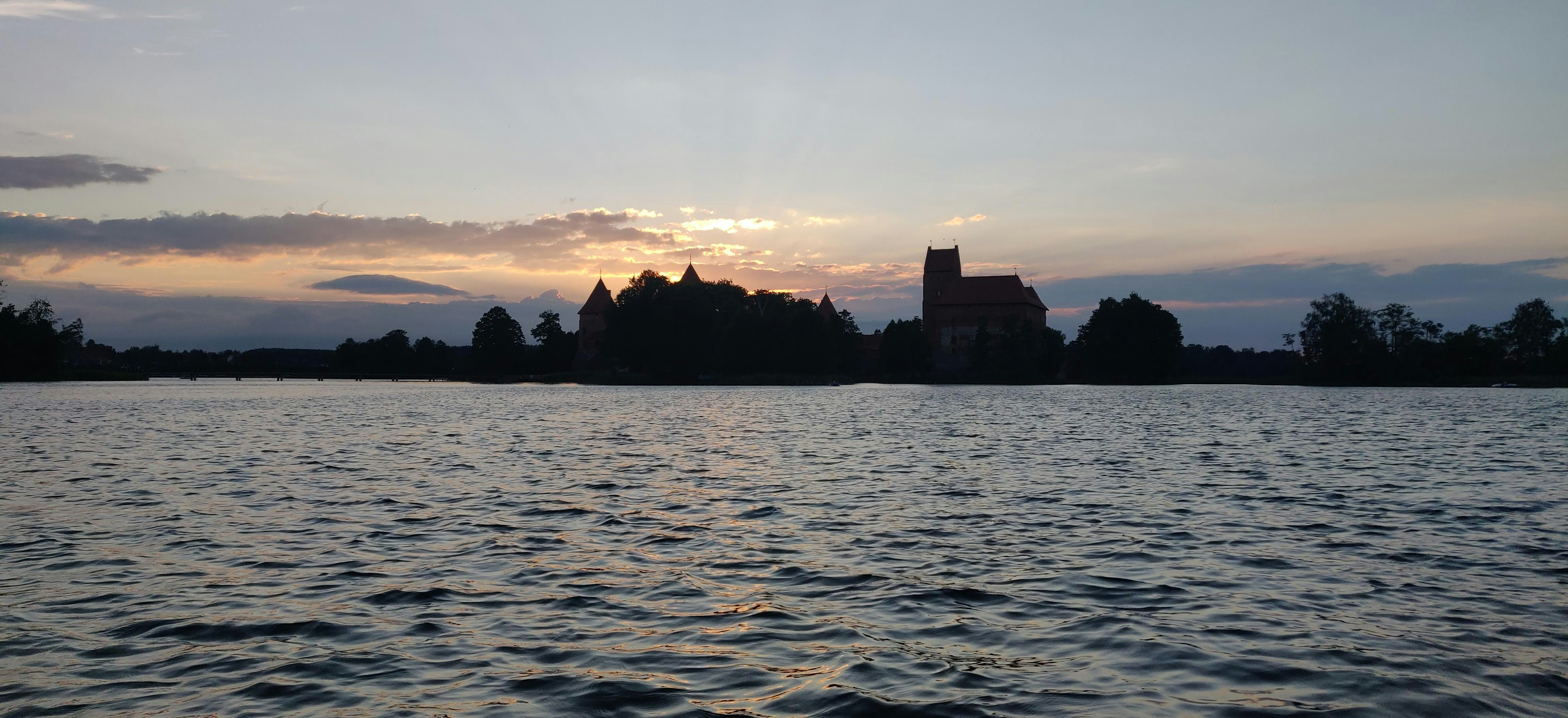 Sunset over a calm lake with a silhouetted church and trees on the horizon.