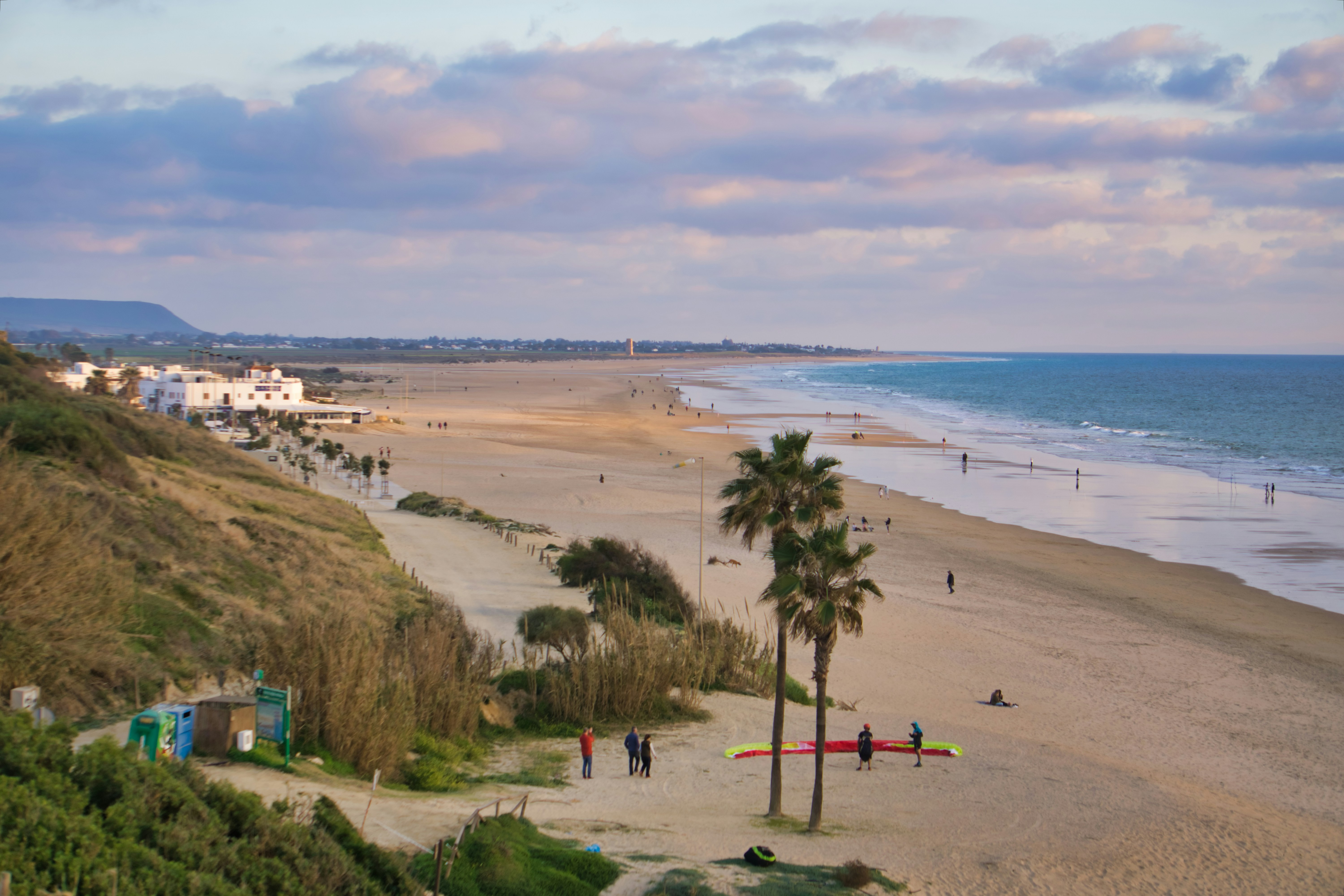 Wide sandy beach in Andalusia with gentle waves and scattered beachgoers under a pastel sky.
