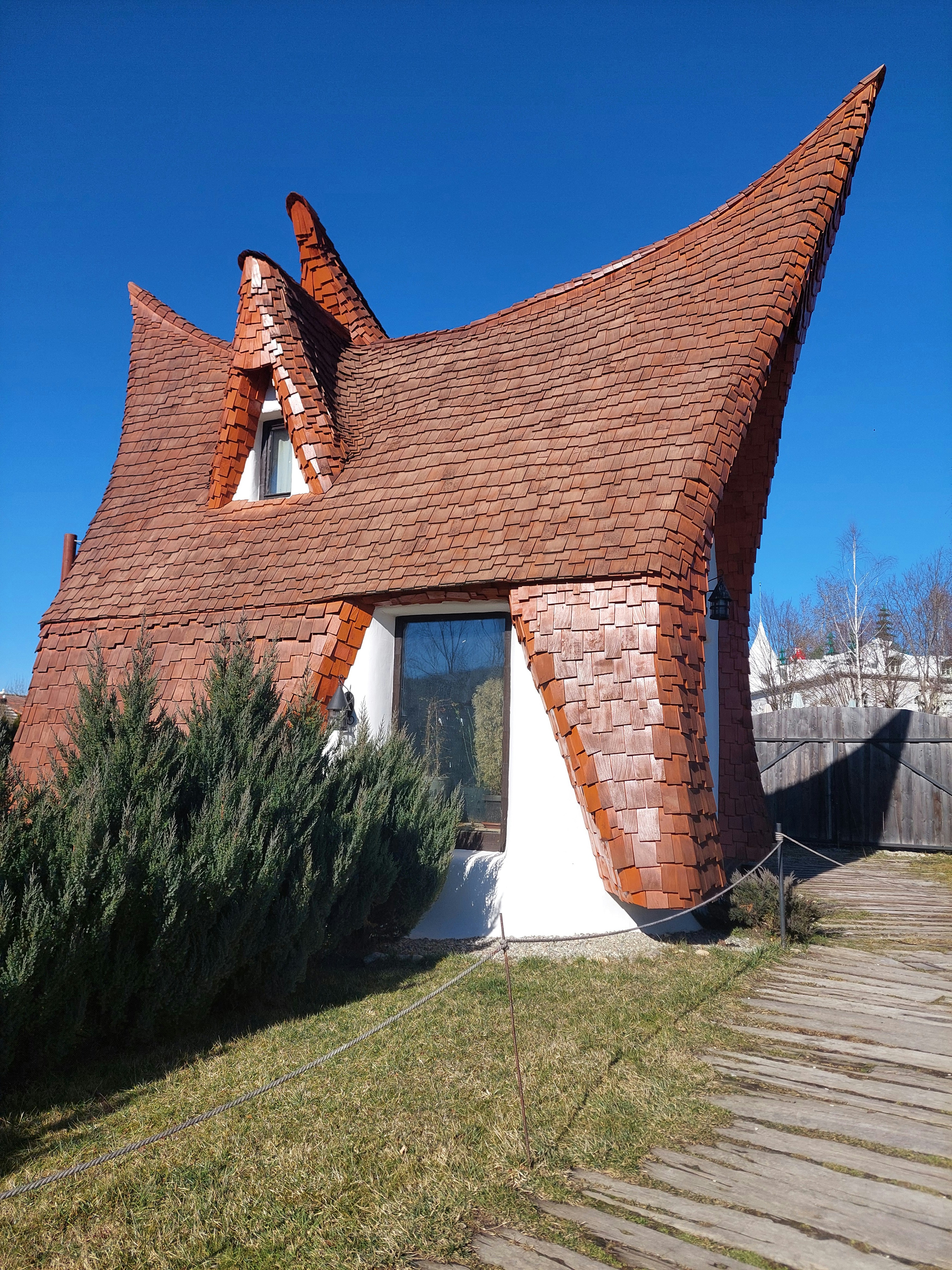 Quirky cottage with a sinuous, brick-clad roof and a small dormer window, flanked by shrubs and a wooden path beneath a clear blue sky.