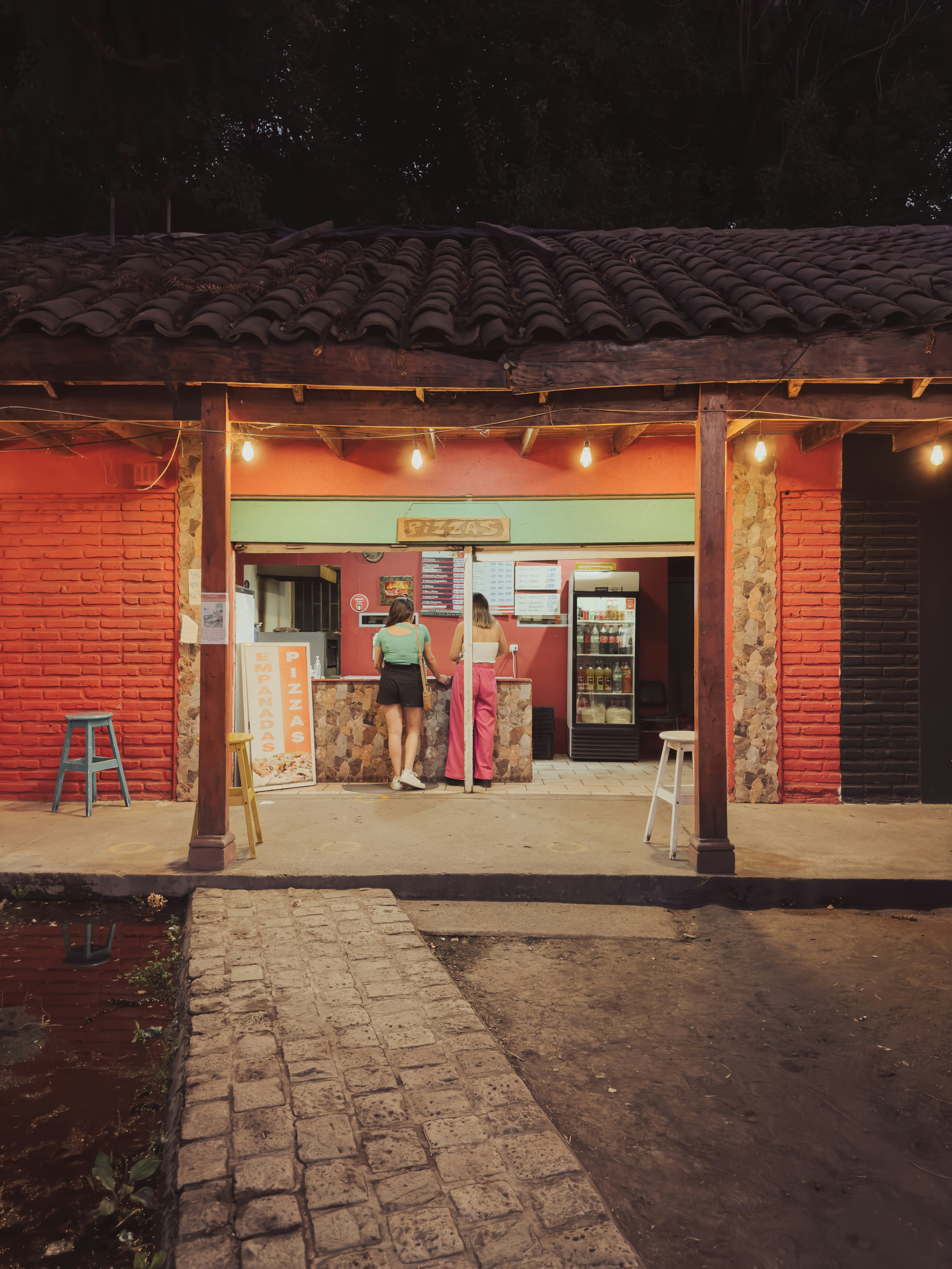 Two people stand at a quaint pizzeria counter adorned with warm lights and rustic decor.