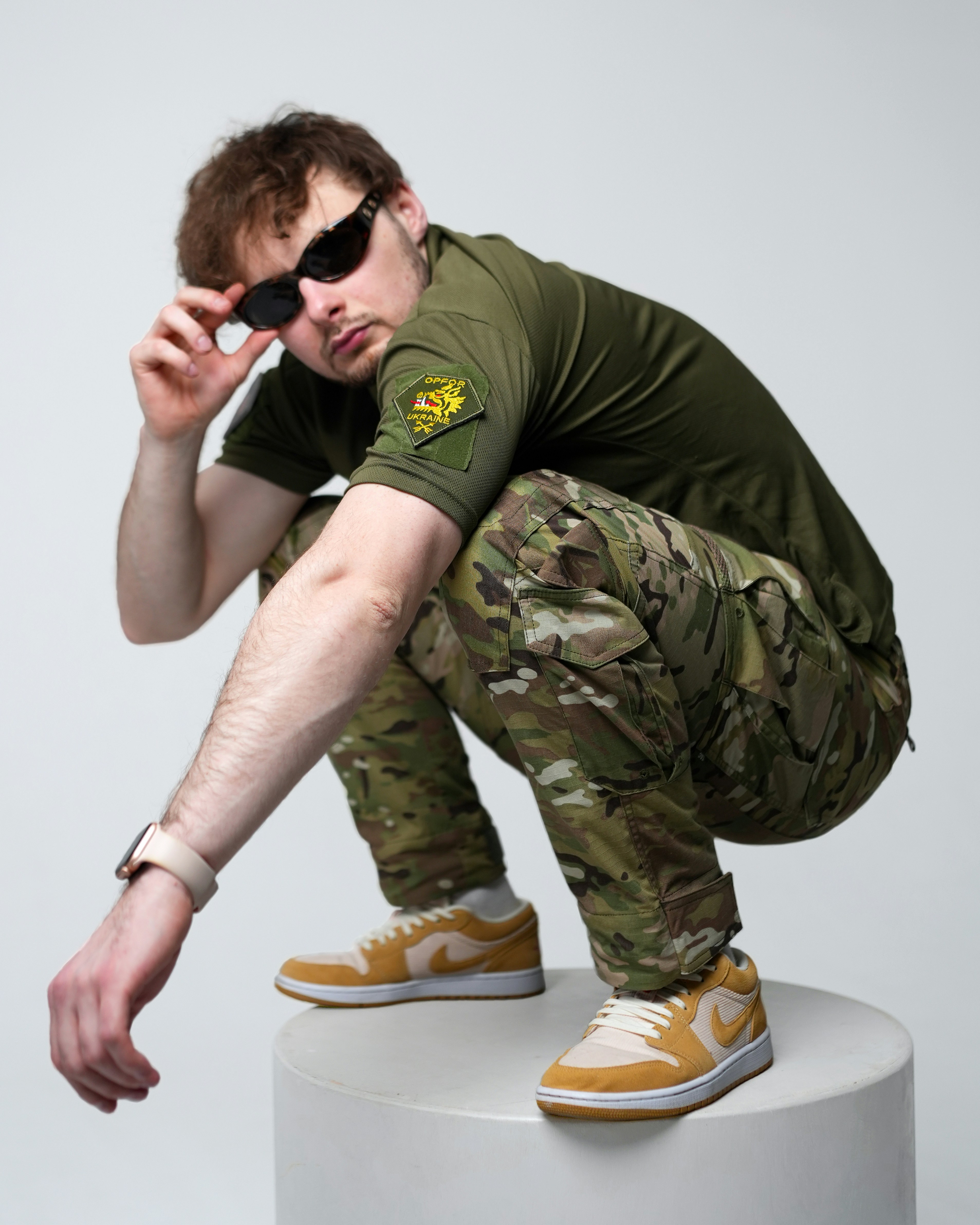 Leather-capped stance: military-casual crouch with sunglasses on a white pedestal
