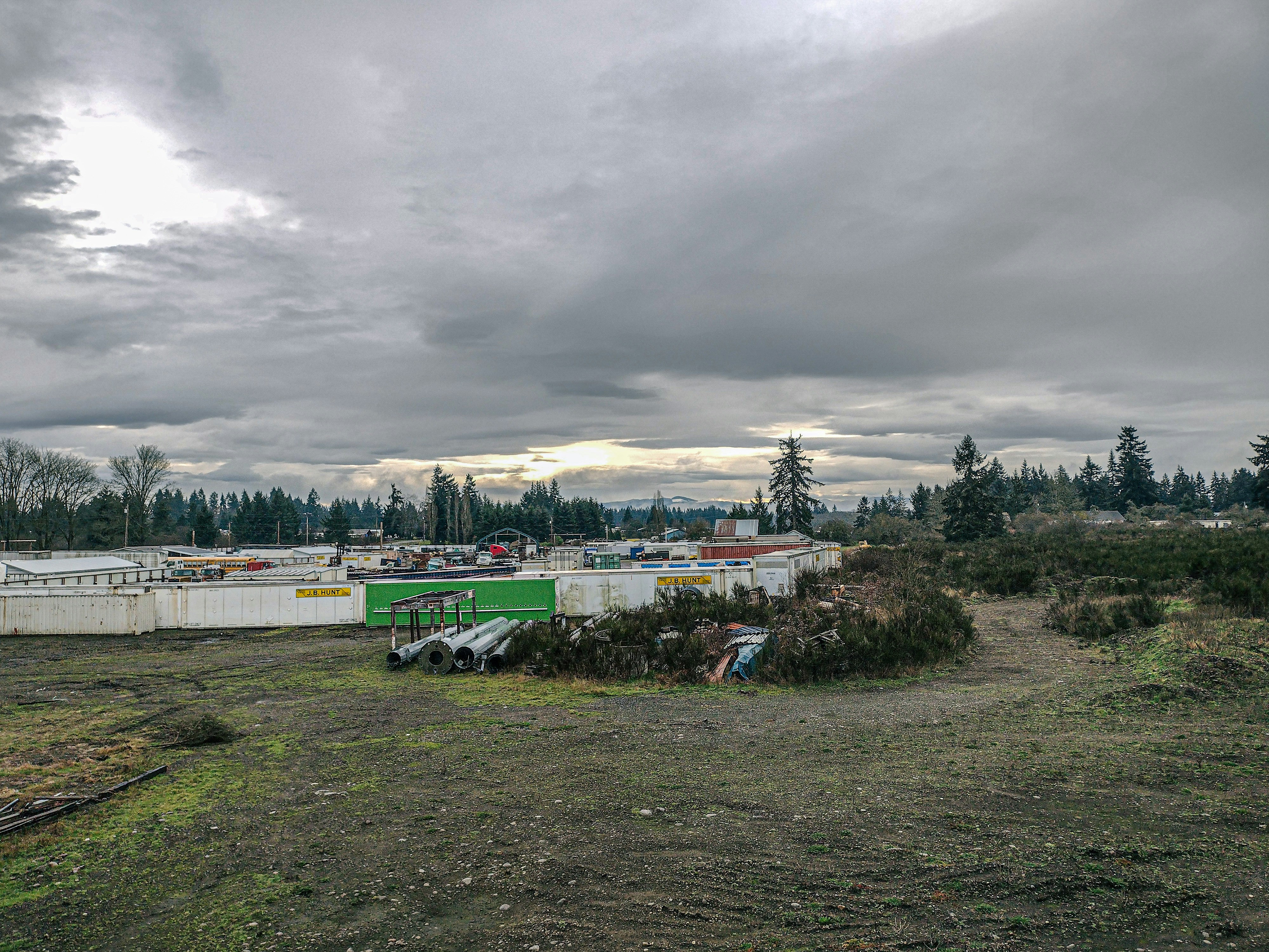 Overcast industrial yard with a row of trailers and scattered equipment stretches across a barren field. A line of green cargo containers draws the eye beneath a heavy, cloud-filled sky.