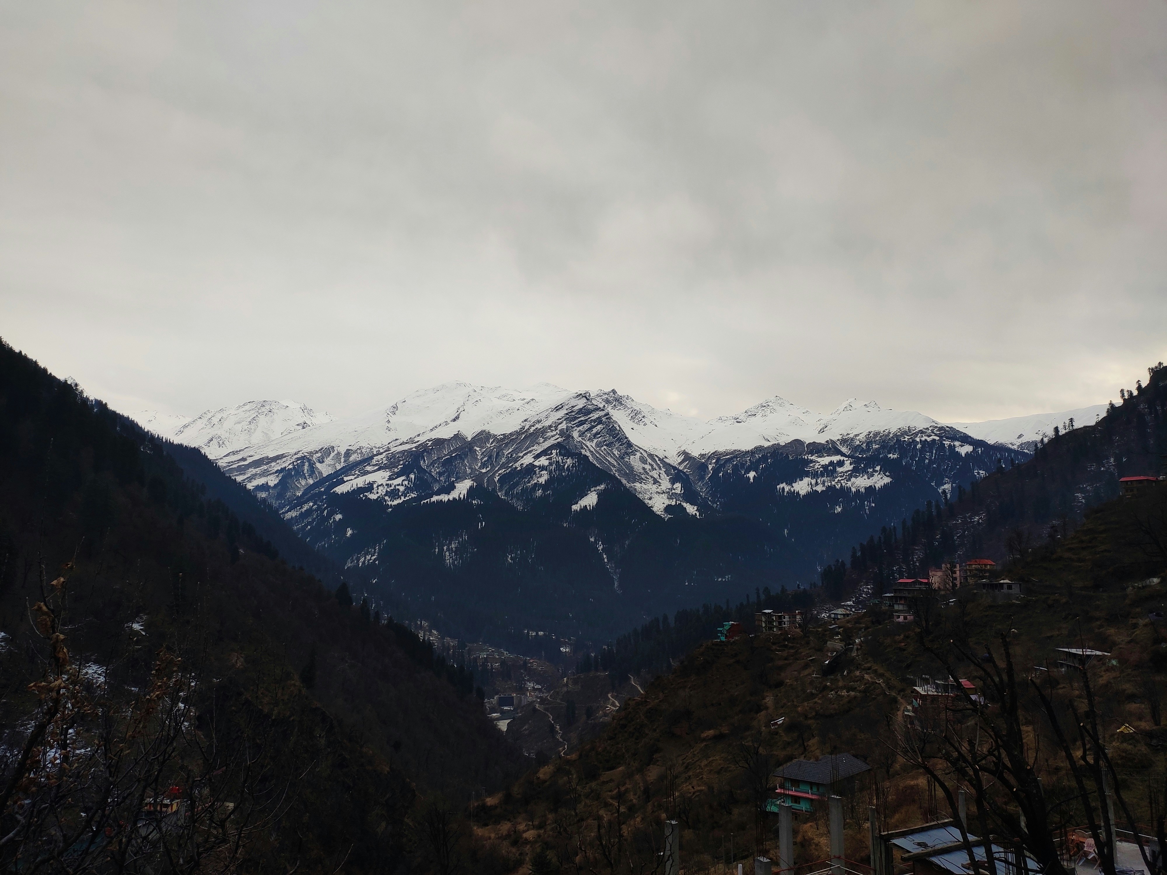 Majestic snowy mountains loom over a tranquil valley, dotted with scattered settlements. The overcast sky adds a serene ambiance.