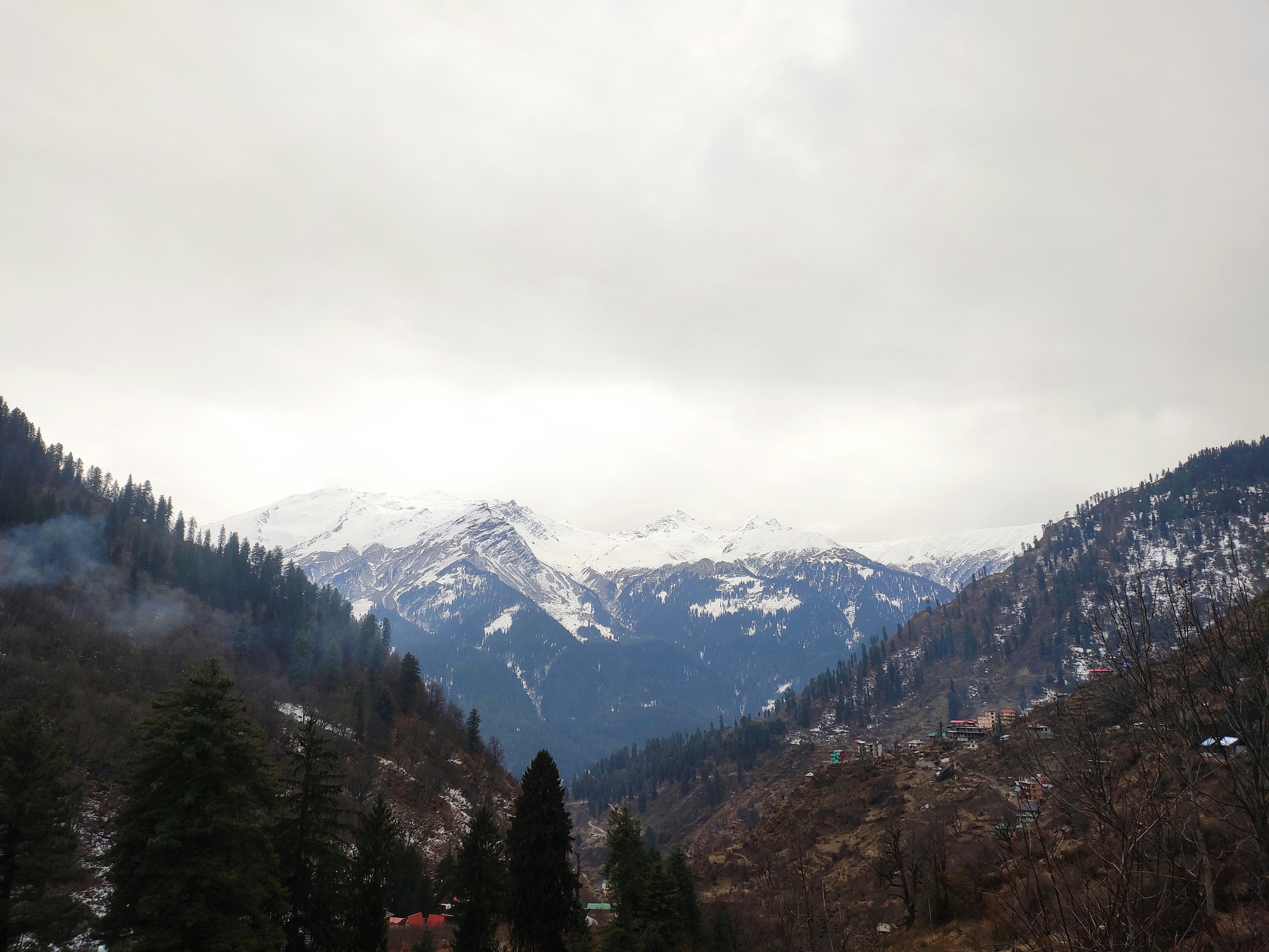 Snow-covered mountains rise above a valley dotted with evergreen trees and scattered buildings.