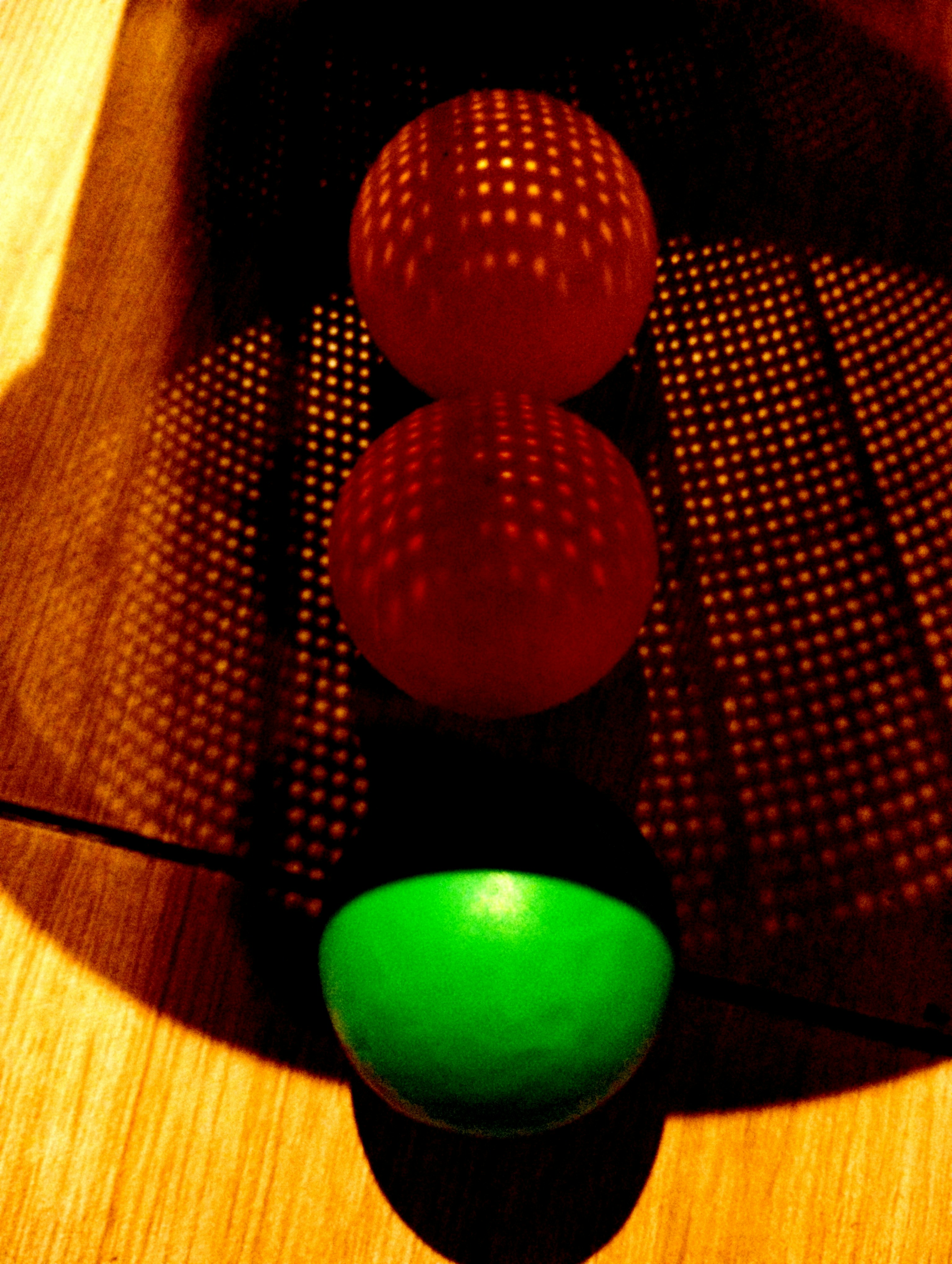 Two red spheres stacked above a green orb sit on a warm wooden surface, with dotted light patterns casting grid-like shadows.