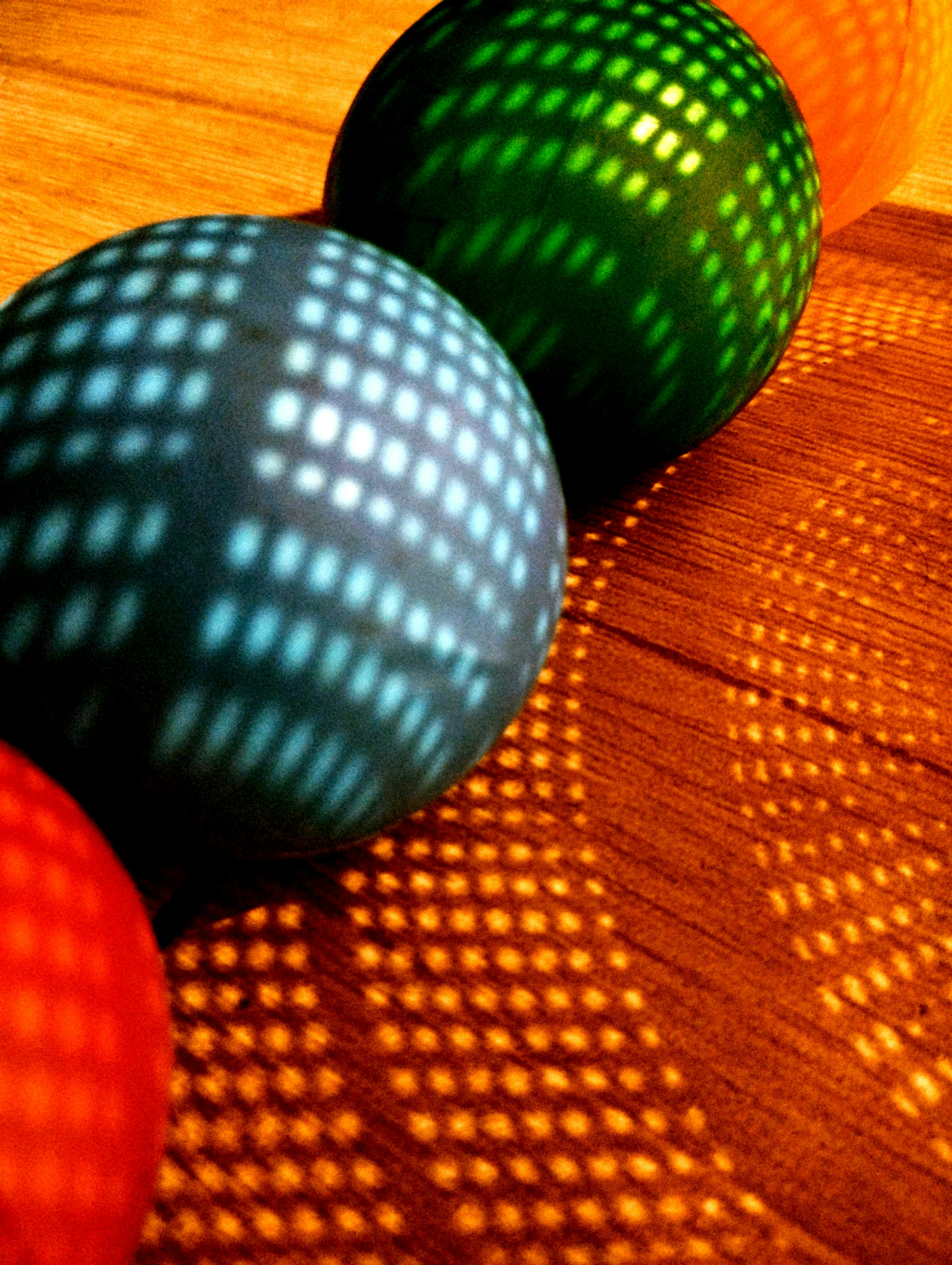 A photograph of colorful balls arranged diagonally on a wooden surface, with dotted light patterns forming a grid. The blue ball dominates the foreground while others recede along the line.