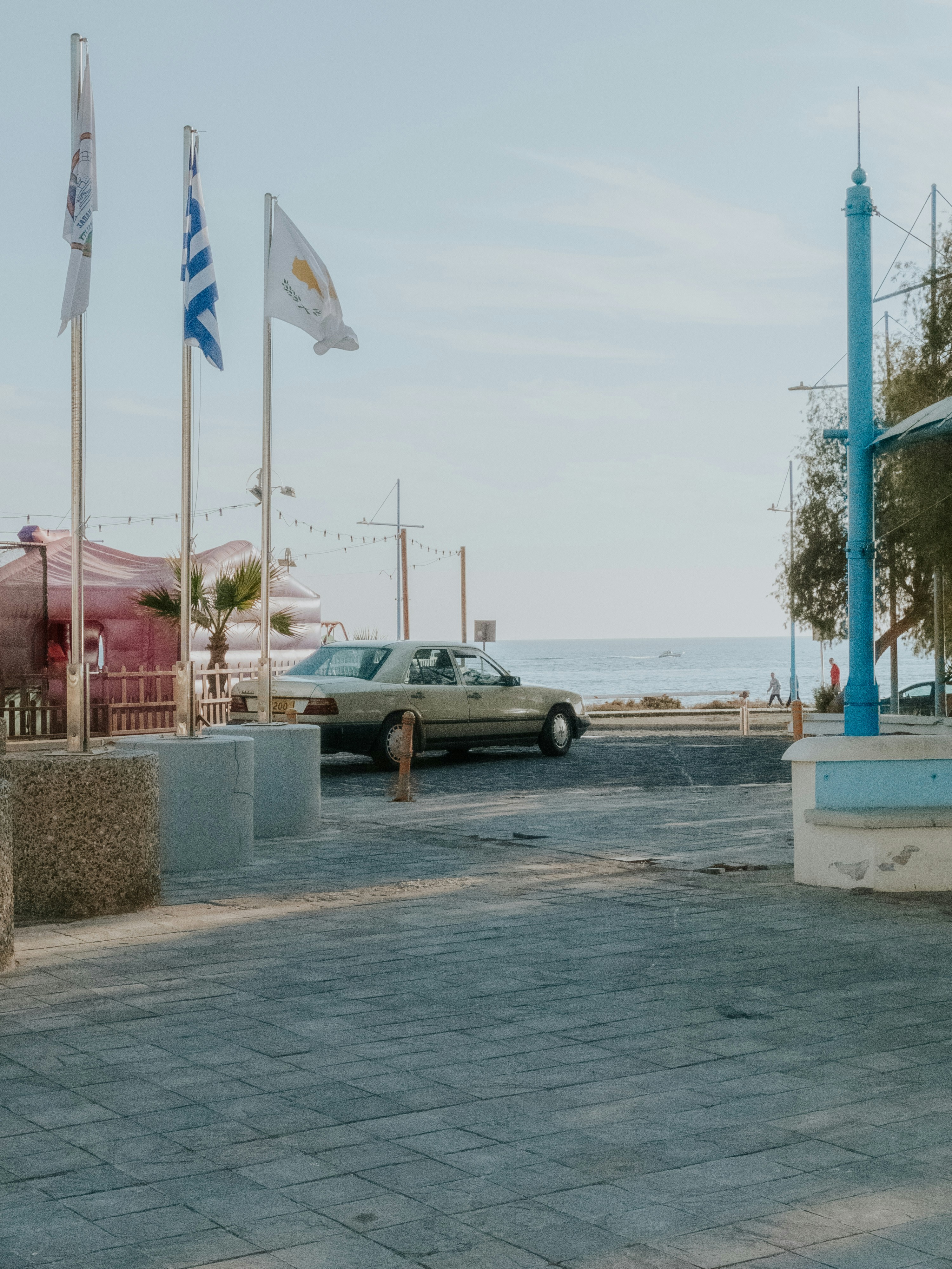 Seaside promenade with a vintage sedan parked by flagpoles and pastel structures. The calm sea and pale sky stretch toward the horizon.