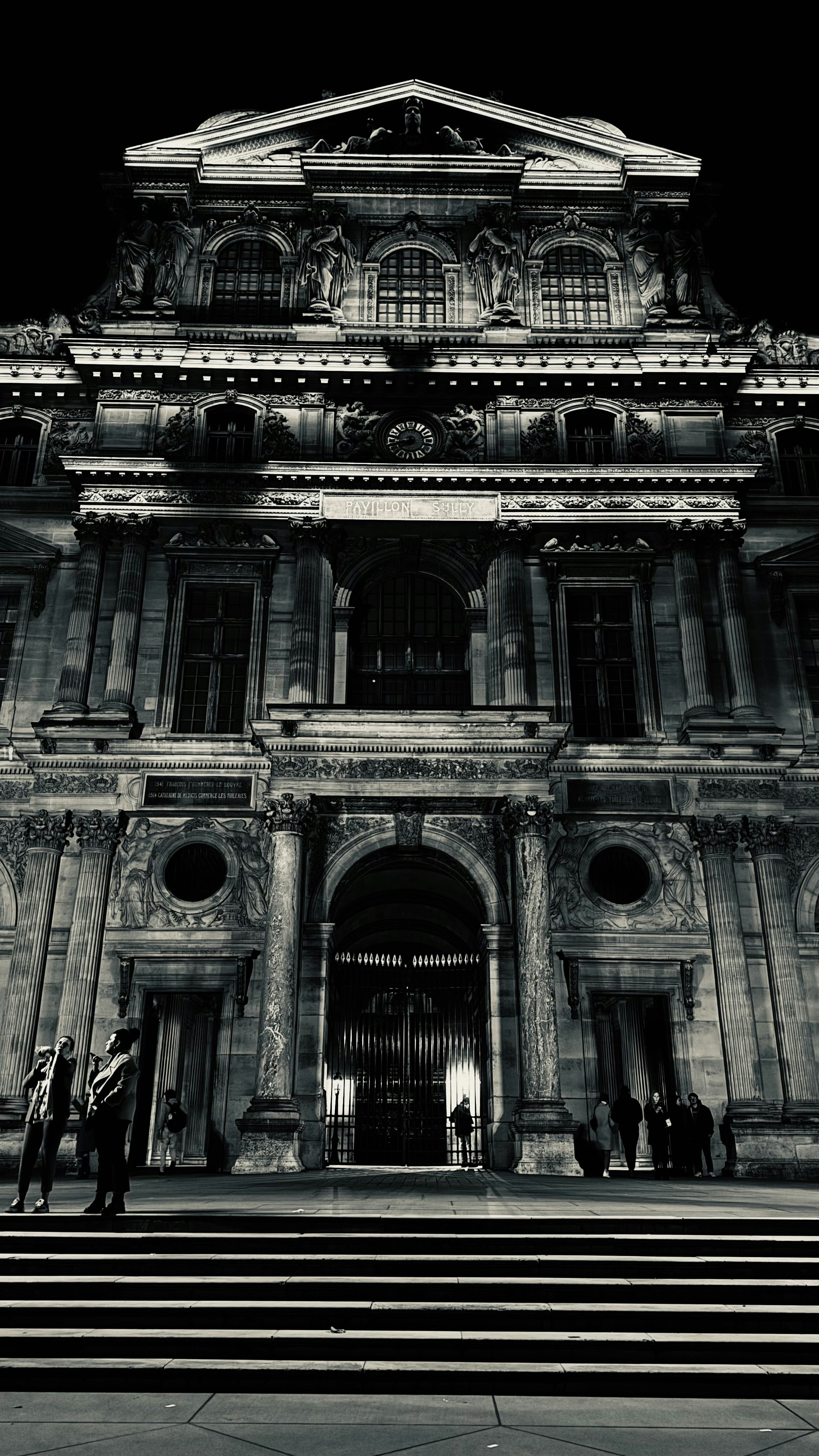 Illuminated facade of the Louvre Museum at night, showcasing intricate architectural details and visitors gathering on the steps.