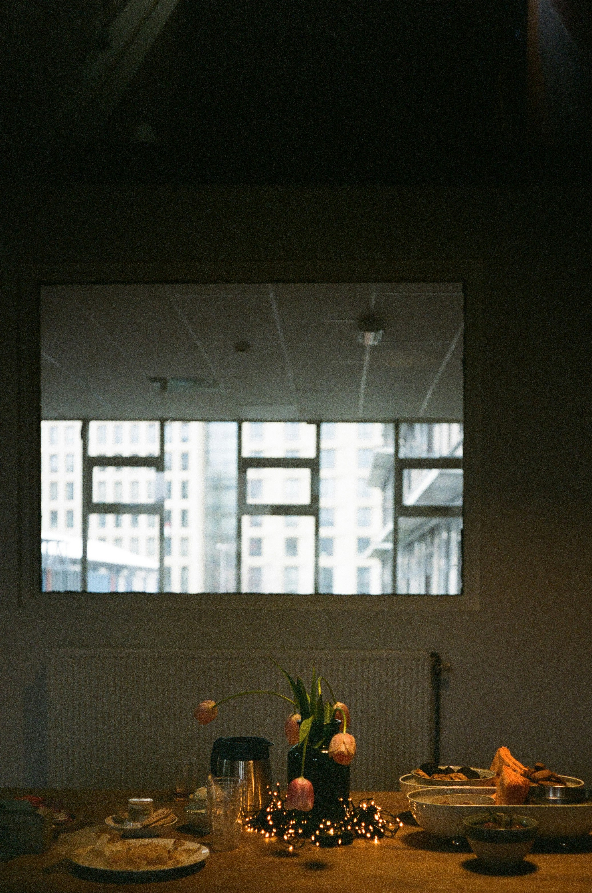 Dimly lit dining table adorned with plates and a string of lights, set against a large window showing urban buildings.