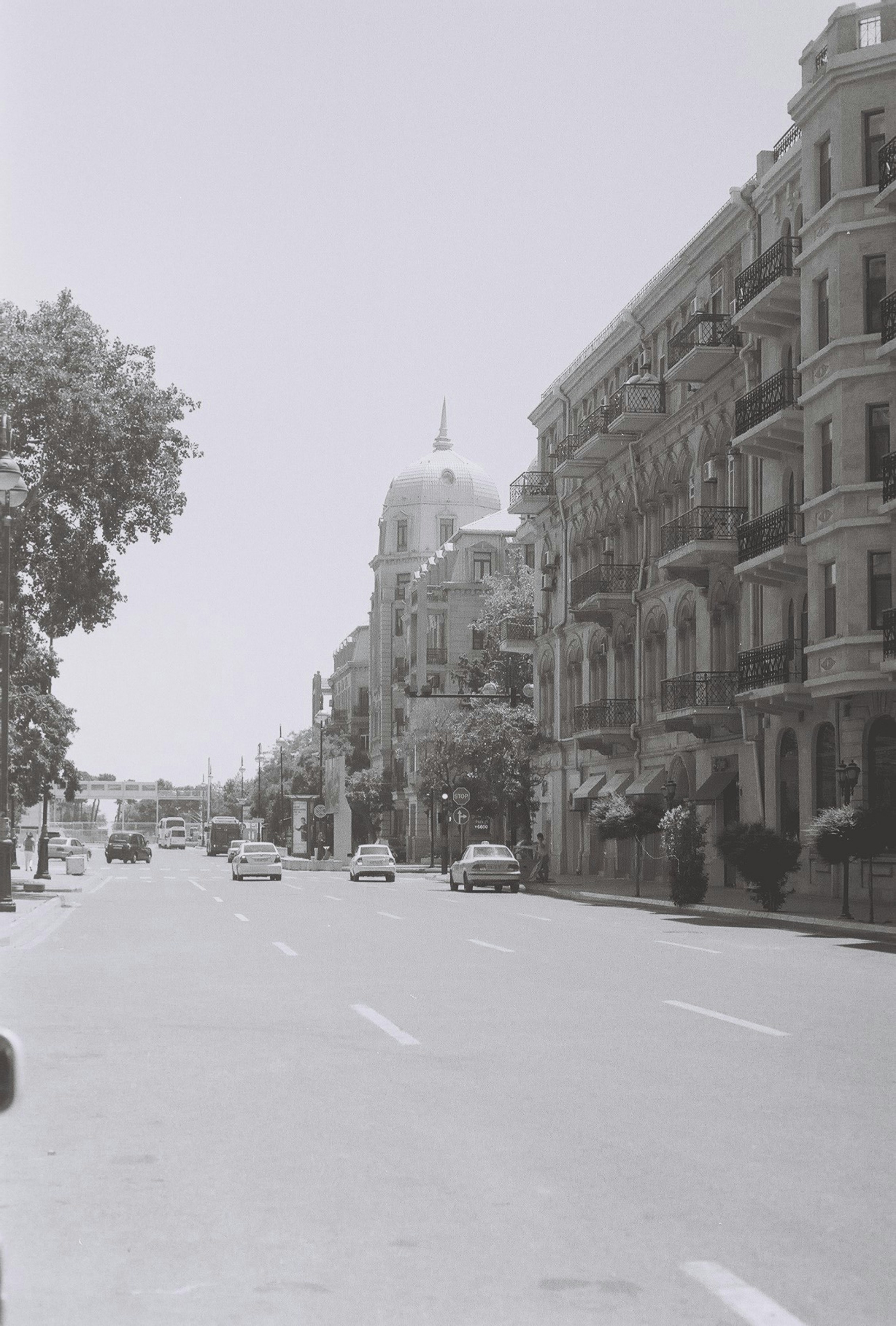 Monochrome street scene photograph showing ornate balconies lining the right-hand buildings and a domed structure on the horizon as cars travel along the wide avenue.