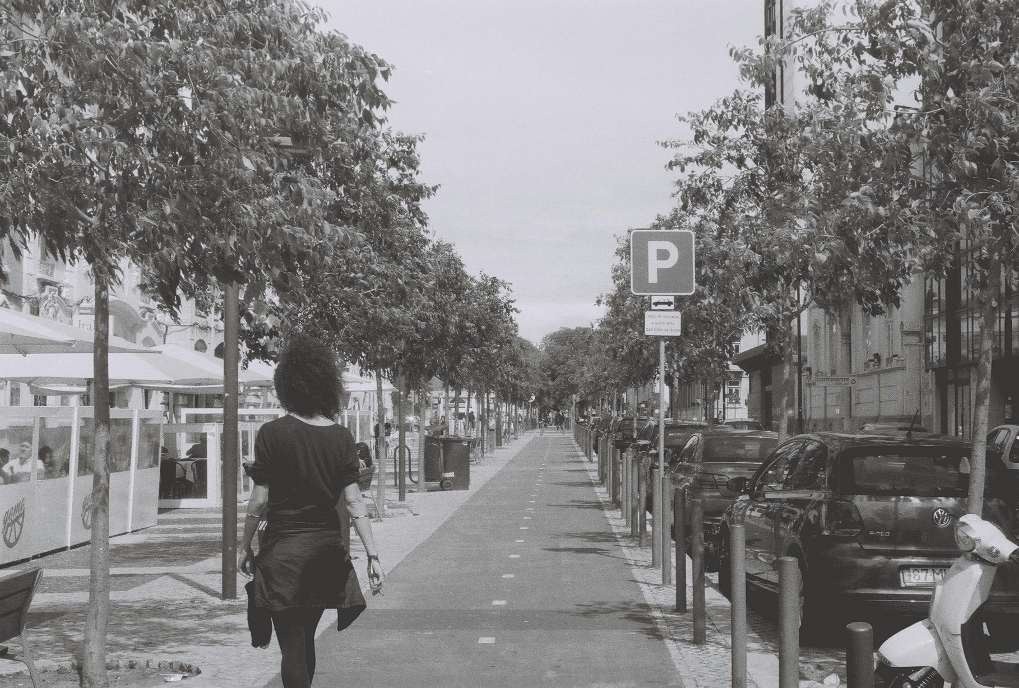 Monochrome street photograph of a pedestrian walking away along a tree-lined avenue with parked cars and a prominent parking sign.