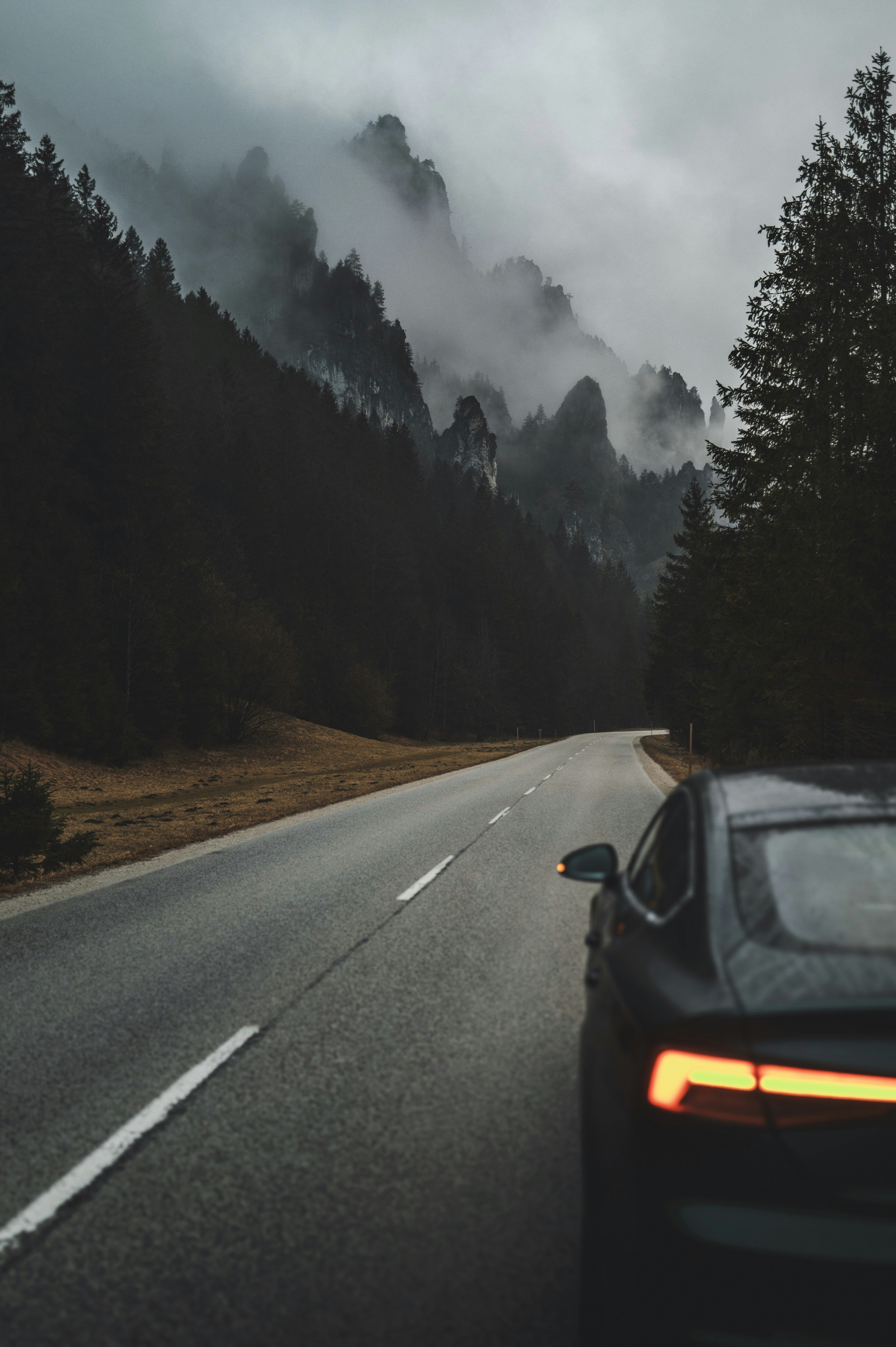 A sleek car parked beside a winding road, framed by towering, mist-covered mountains and dense forests.