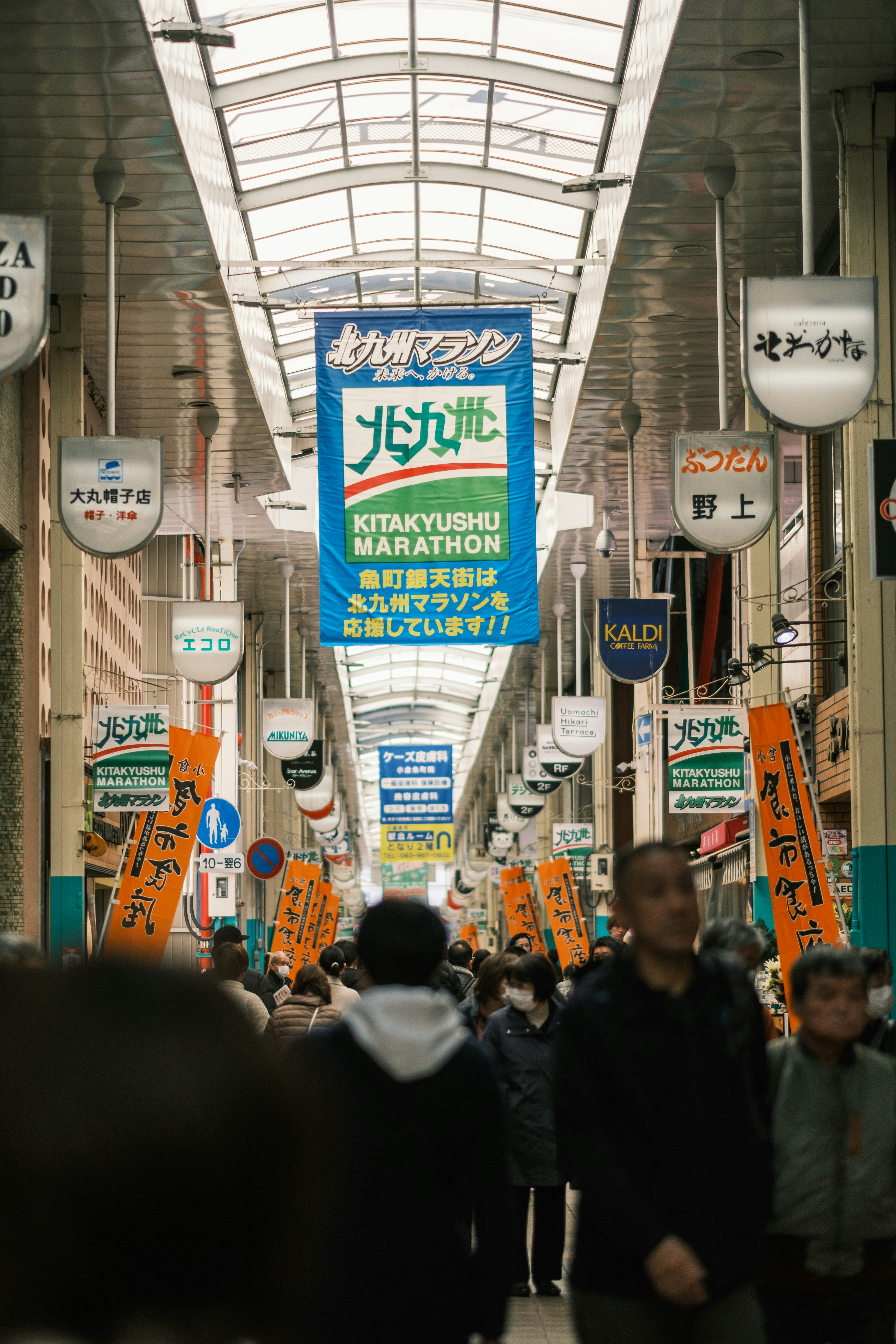 Crowded shopping arcade with colorful banners and signs hanging from the ceiling, bustling with people.