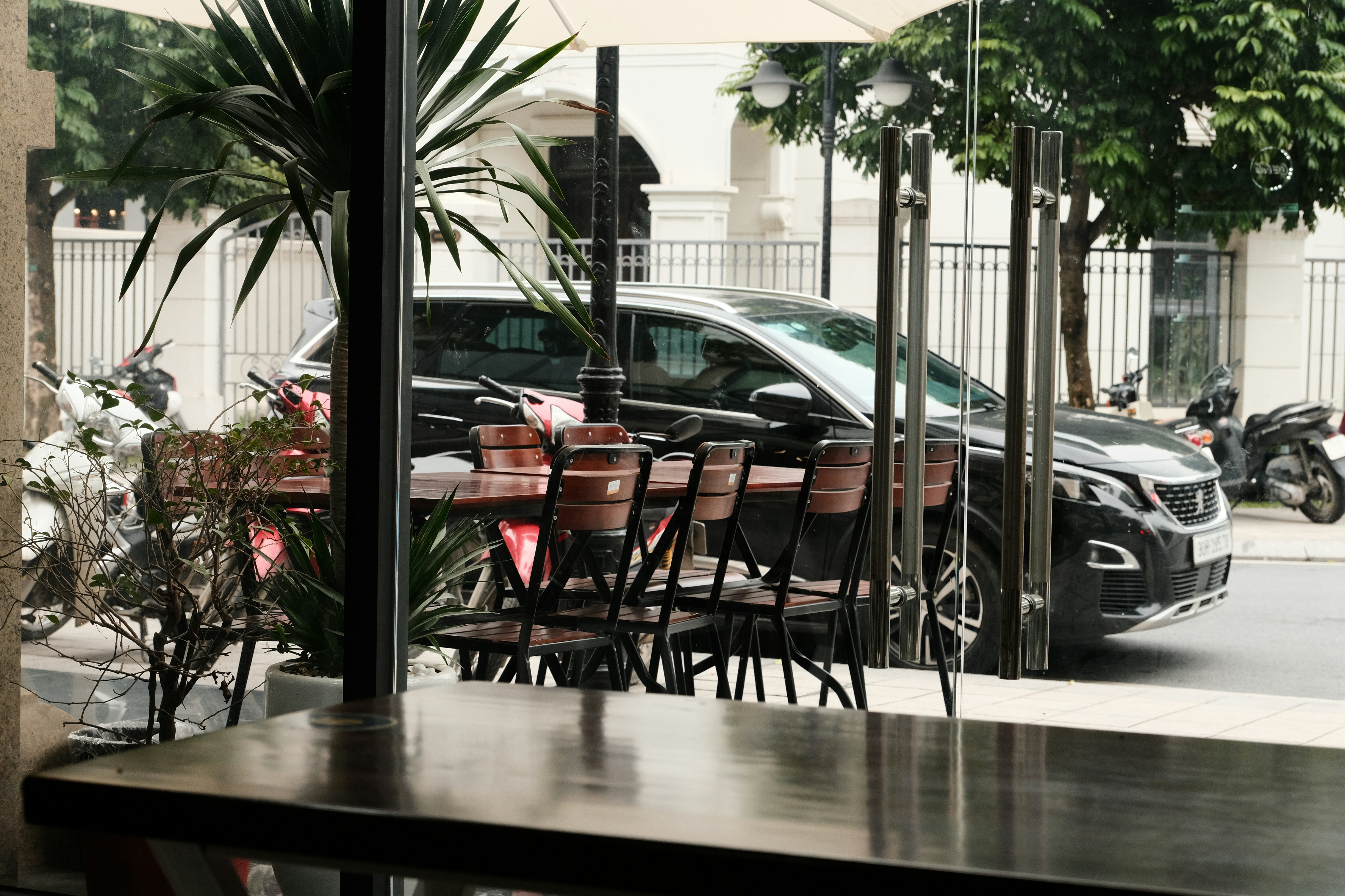 Outdoor seating area with wooden chairs and tables seen through a glass window, with a black car and street in the background.