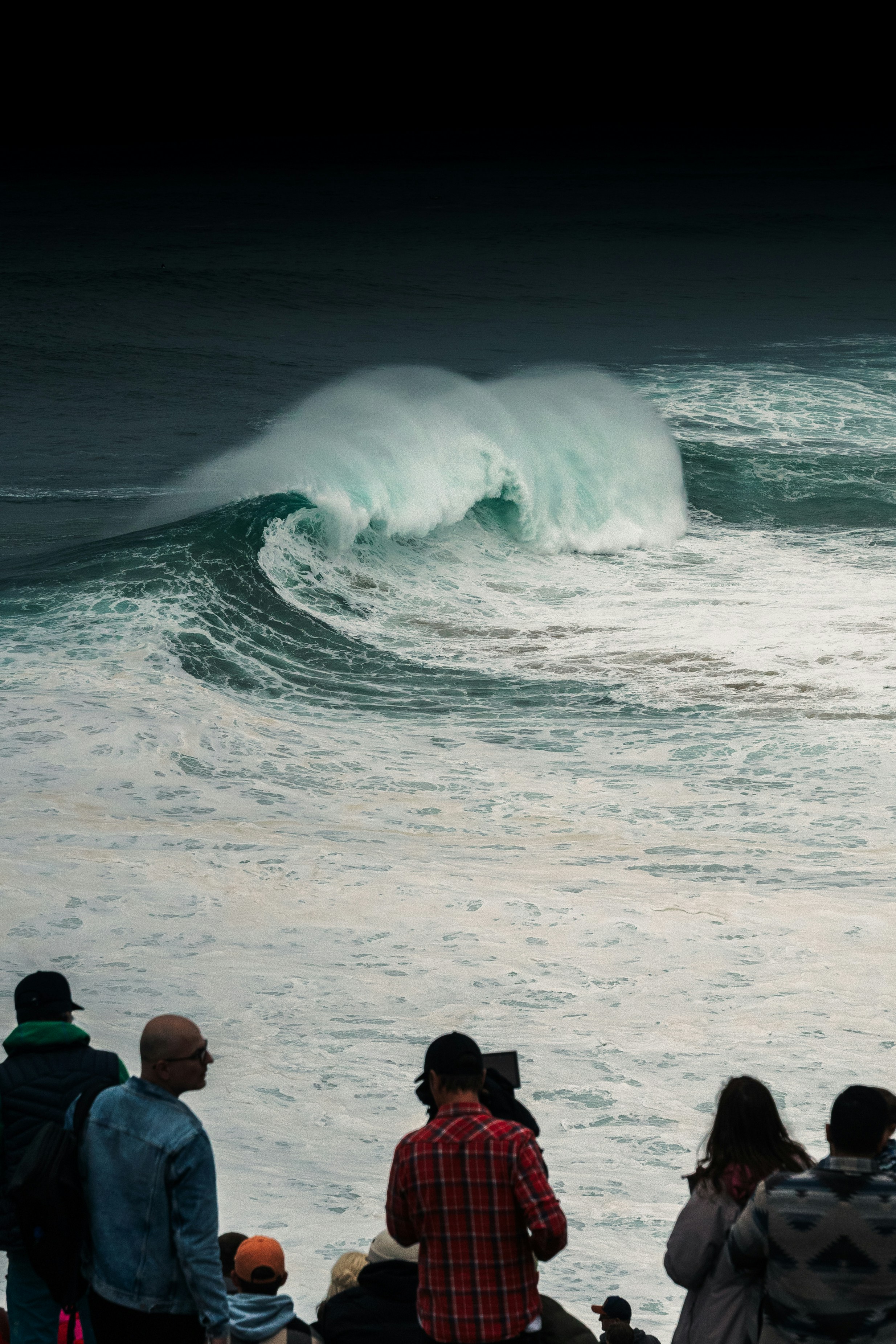 Turquoise Crest at the Shore: A Riding Wave Framed by Onlookers under a Moody Sky
