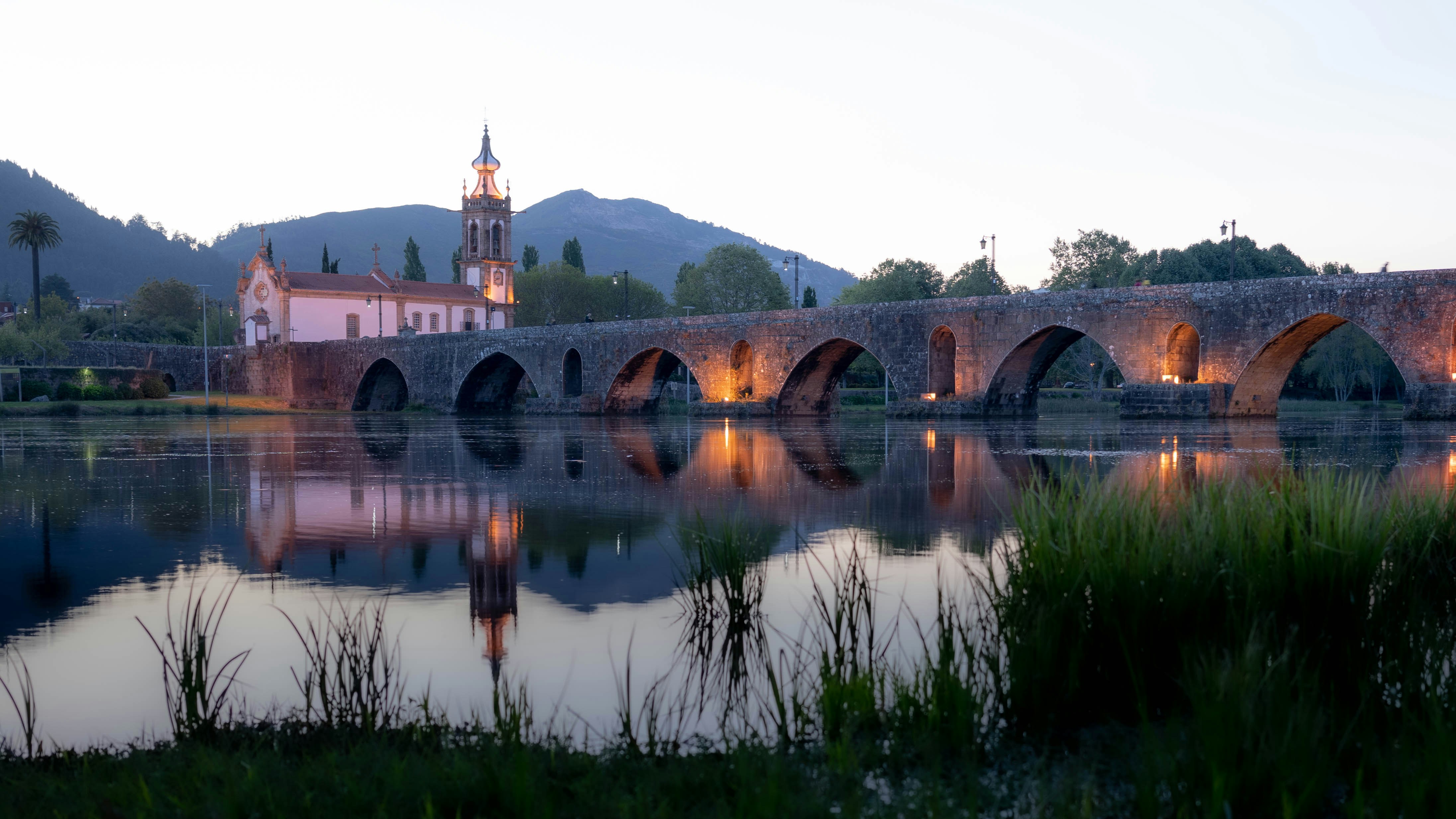 A historic stone bridge arches gracefully over calm waters, reflecting the silhouette of a quaint church set against the soft hues of a sunset sky. This peaceful scene captures the essence of timeless beauty.