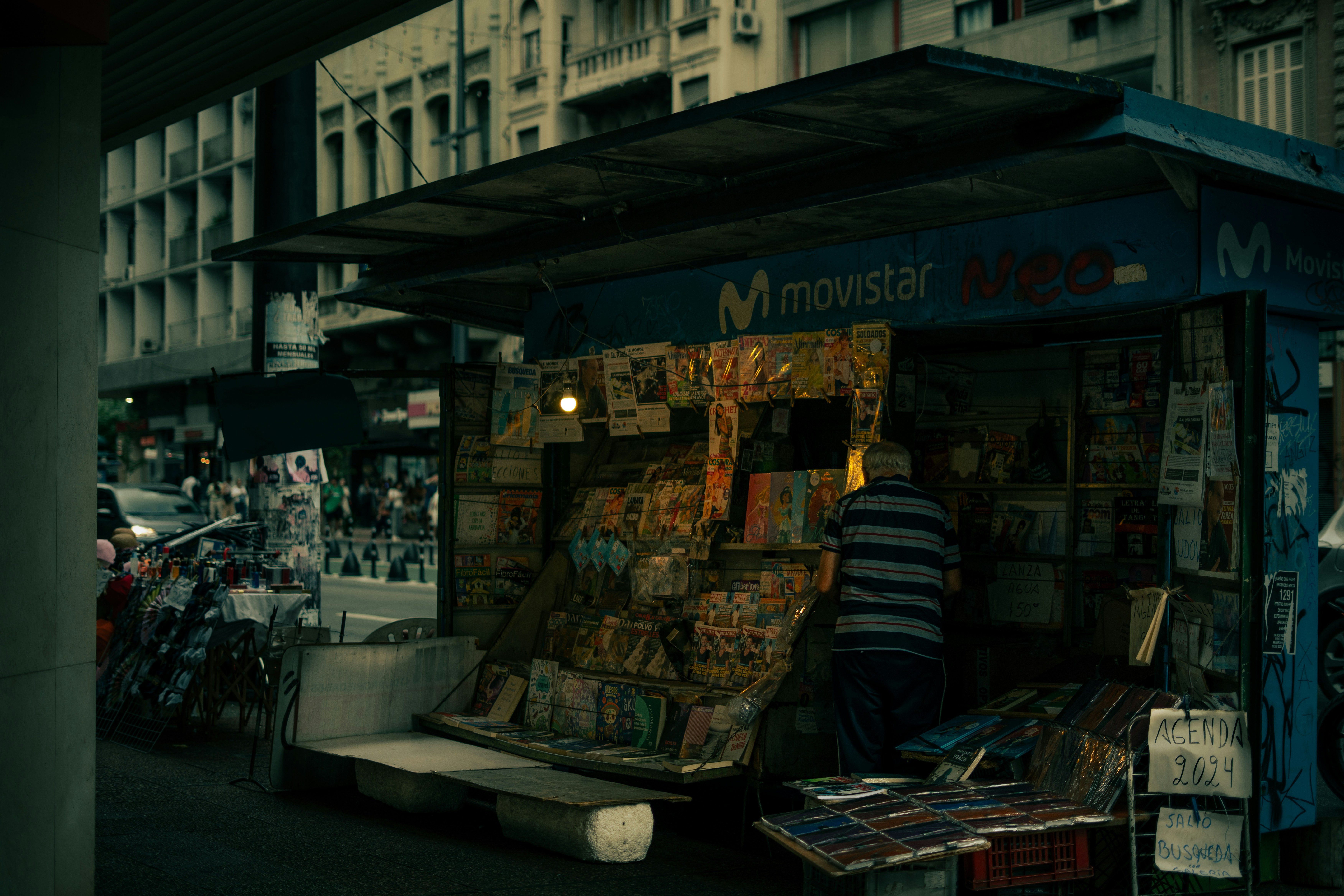 Dimly lit newsstand with illuminated interior contrasts against urban backdrop.