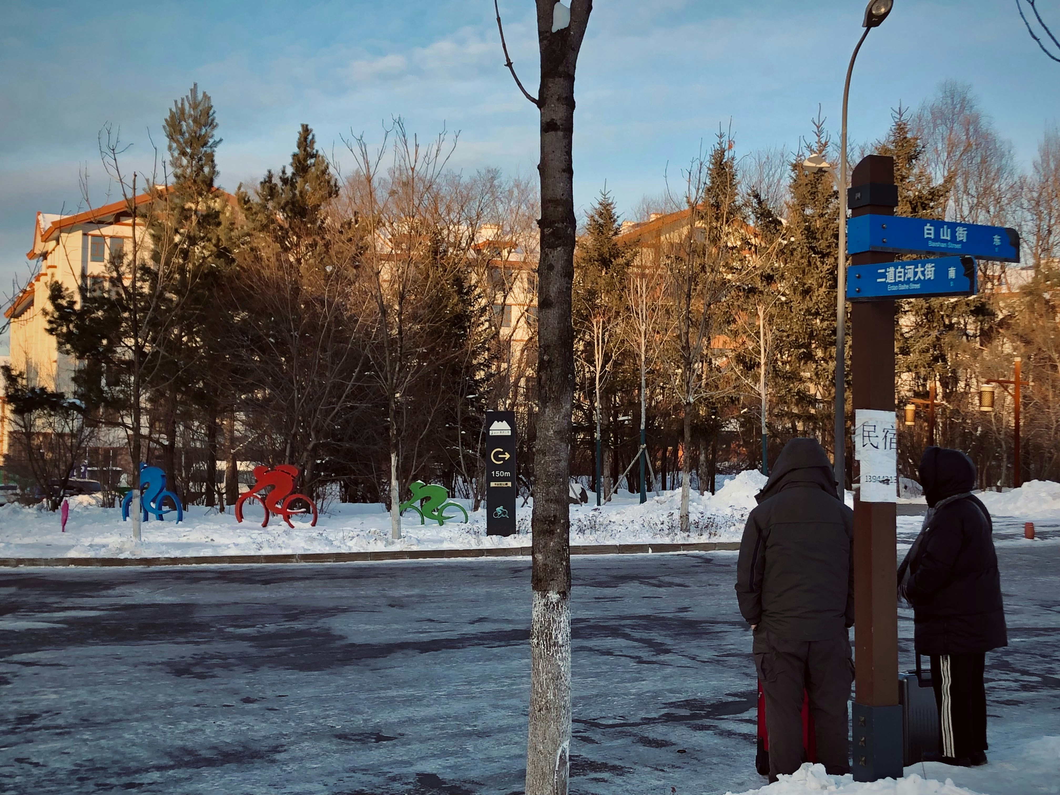 Snowy Street Corridor: Blue Signpost, Two Figures, and a Row of Colorful Reindeer