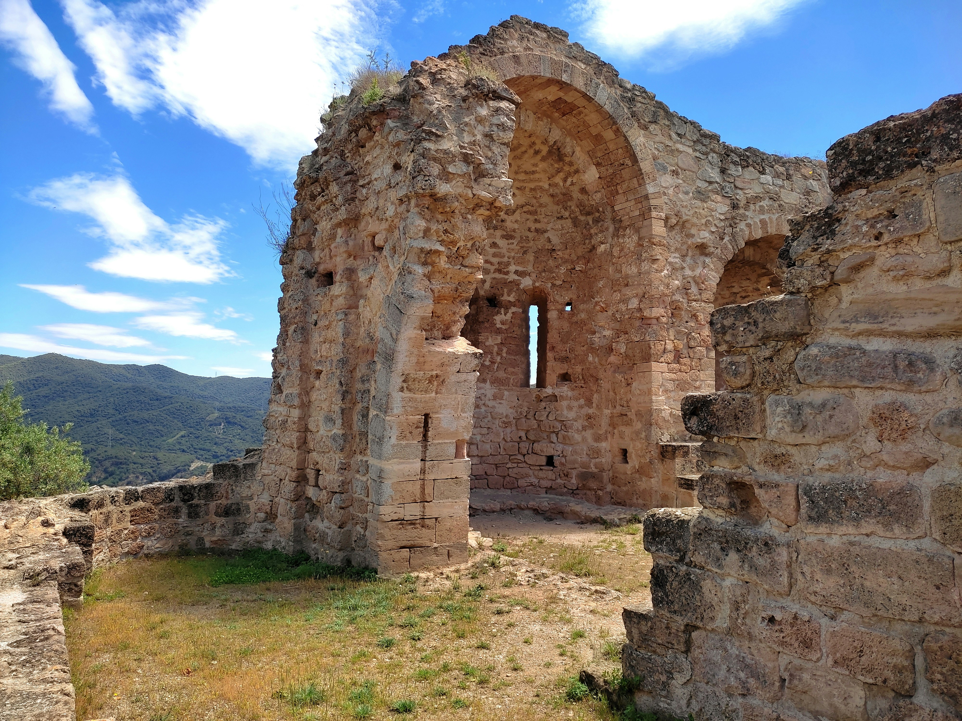 Ancient stone arches of a hilltop ruin rise against a bright blue sky, with distant mountains on the horizon. Weathered brick walls and grassy foreground frame the scene.