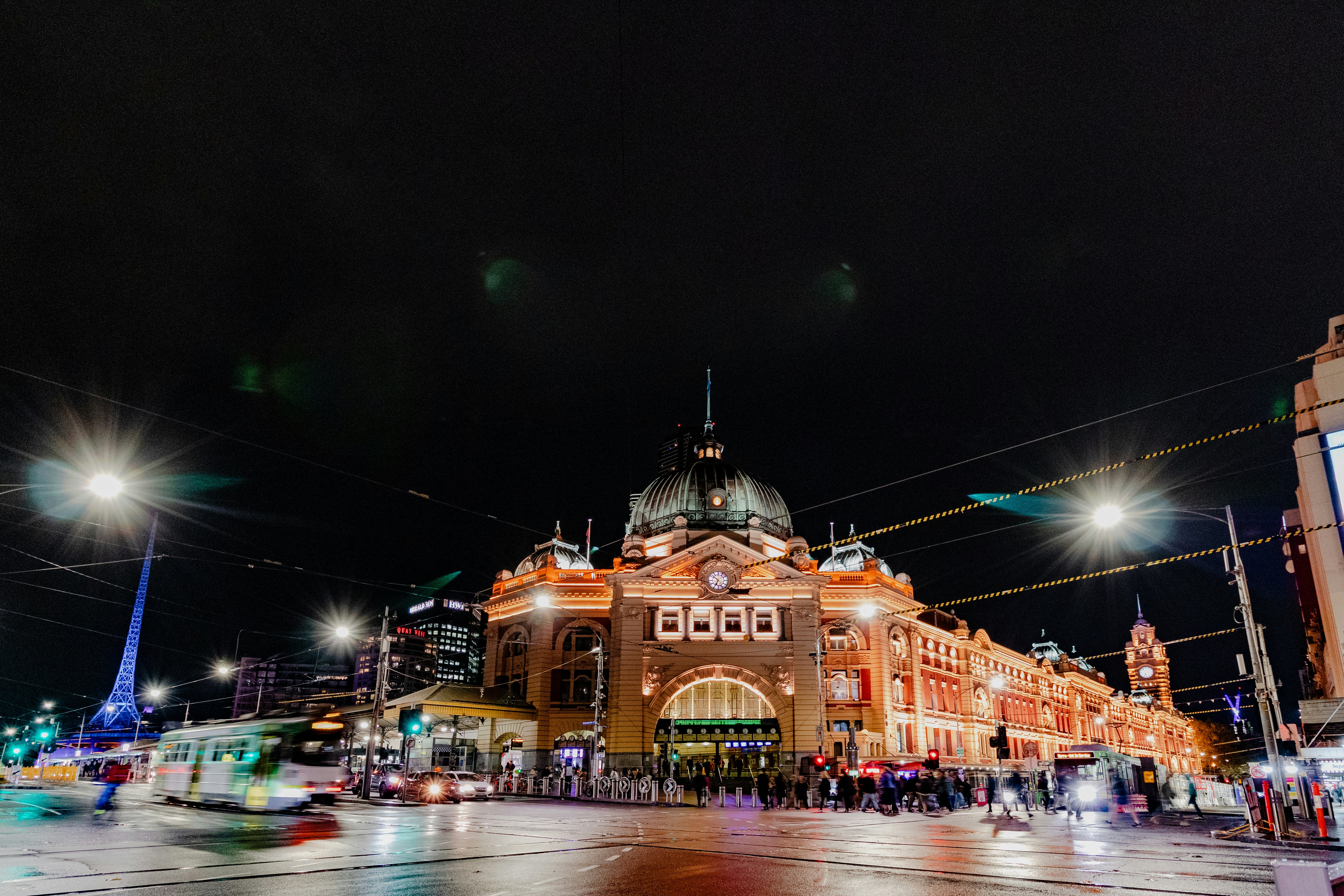Historic train station glowing with vibrant lights against a night sky, with blurred trams and bustling pedestrians.