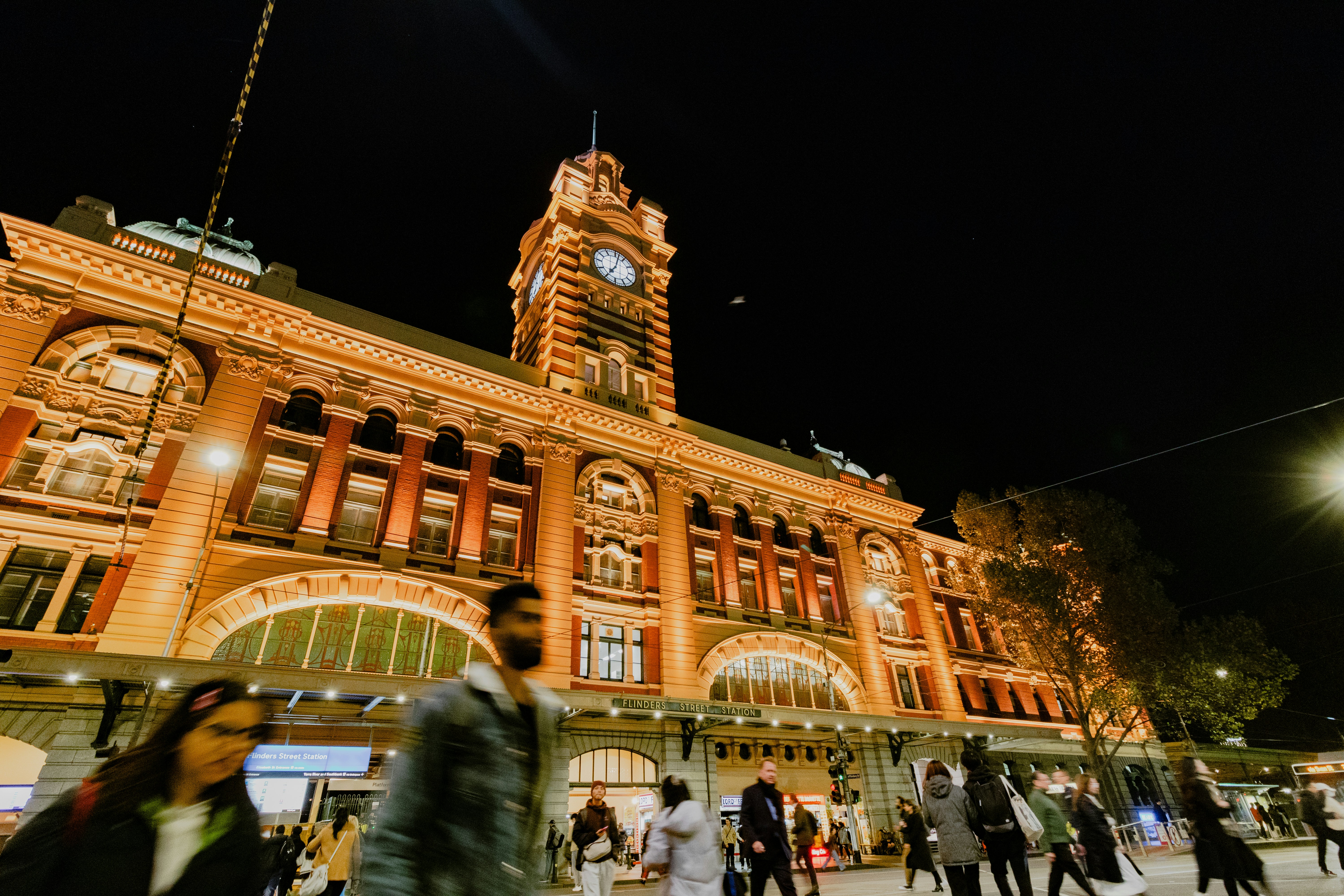 Illuminated Flinders Street Station with blurred pedestrians in the foreground under a clear night sky.