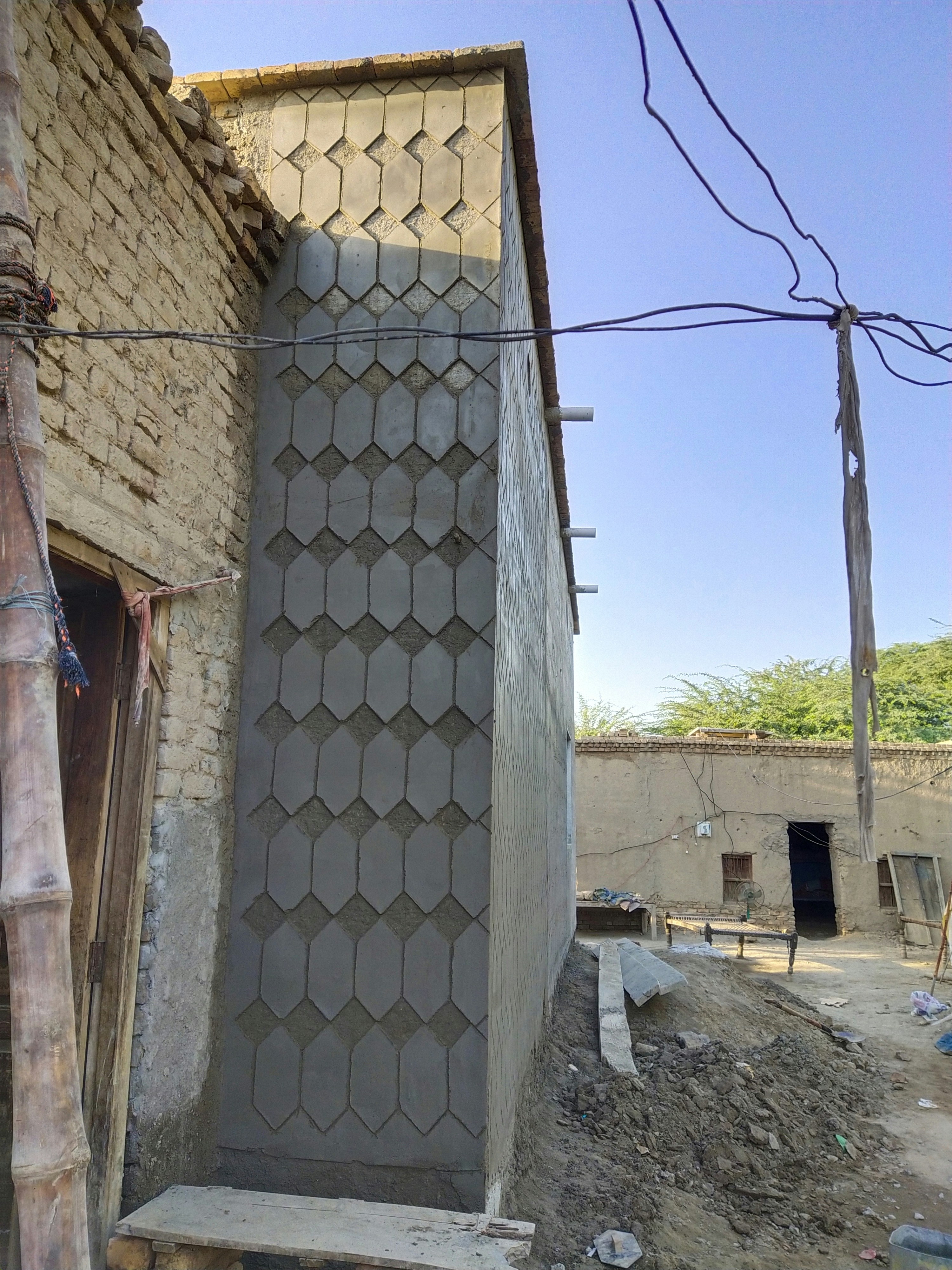 Daylight photograph of a tall hexagonal-tiled wall rising along a dusty construction yard, with overhead wires and rough ground.